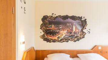 Quarto de hotel com cama, cabeceira de madeira, luminária fixada na parede e um mural com vista para a cidade.