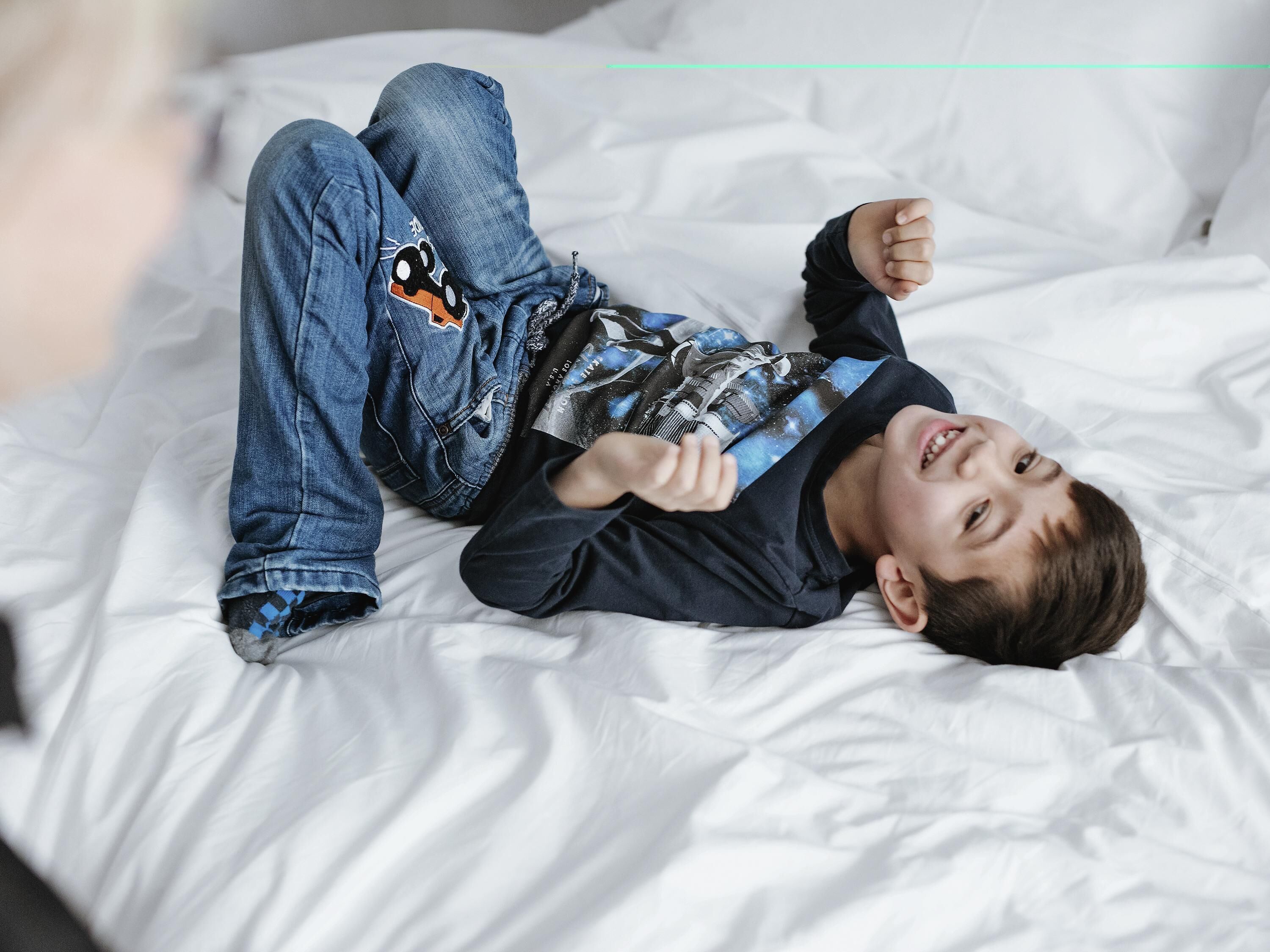 Uma pessoa deitada em uma cama com roupa de cama branca, vestindo uma camisa escura e jeans.