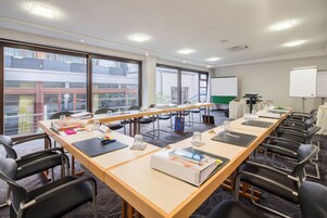 Sala de conferências com mesas compridas, cadeiras, um quadro branco e vista para prédios através de grandes janelas.