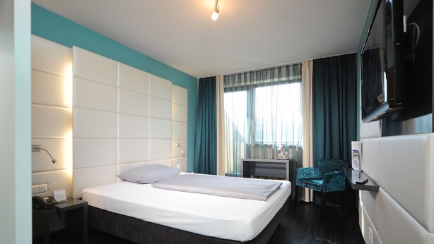 Quarto de hotel com uma cama grande, uma escrivaninha, uma cadeira, uma televisão e uma janela com cortinas.