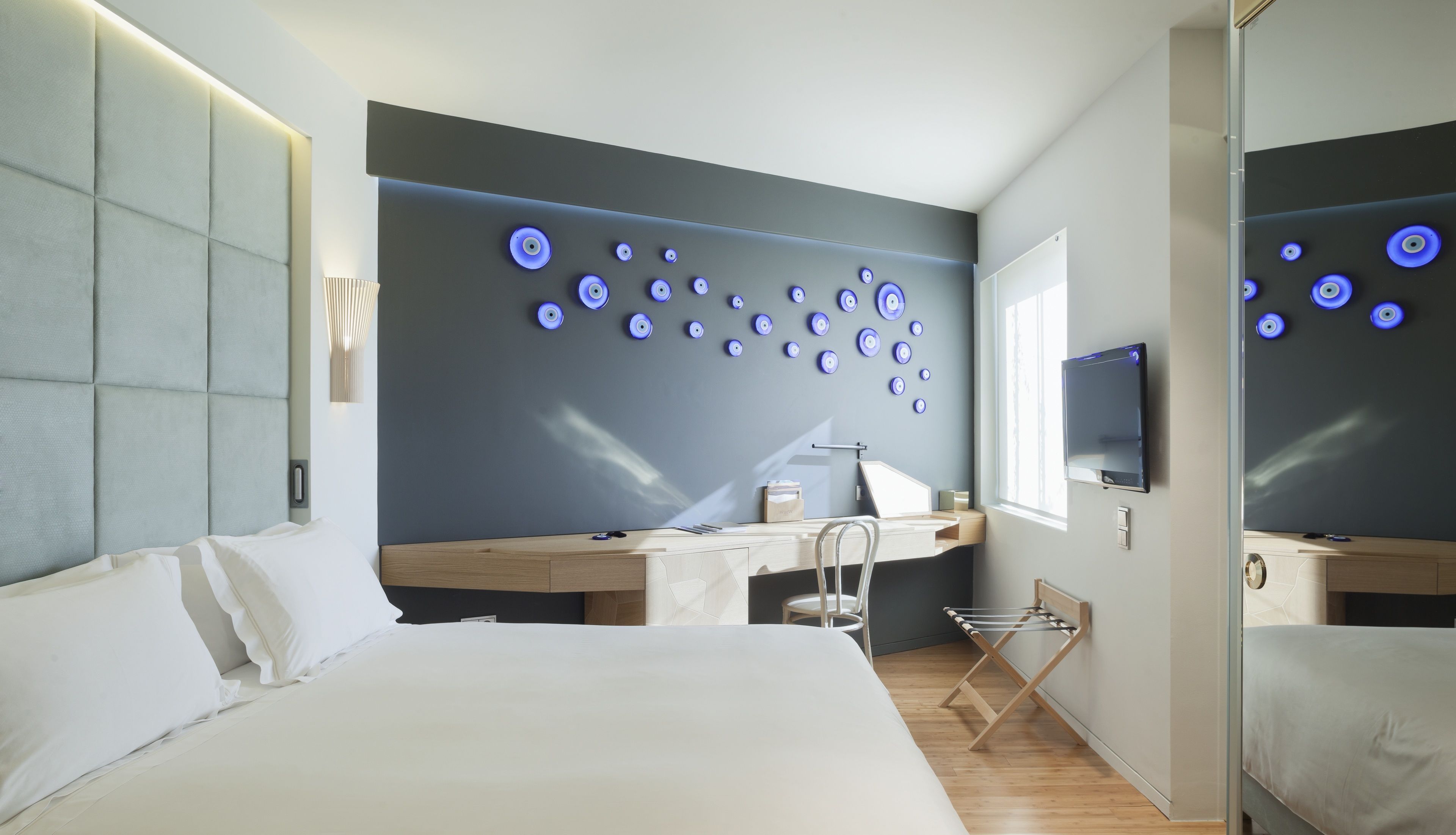 Quarto de hotel moderno com uma cama grande, uma escrivaninha com cadeira, uma televisão e uma parede com luminárias circulares.