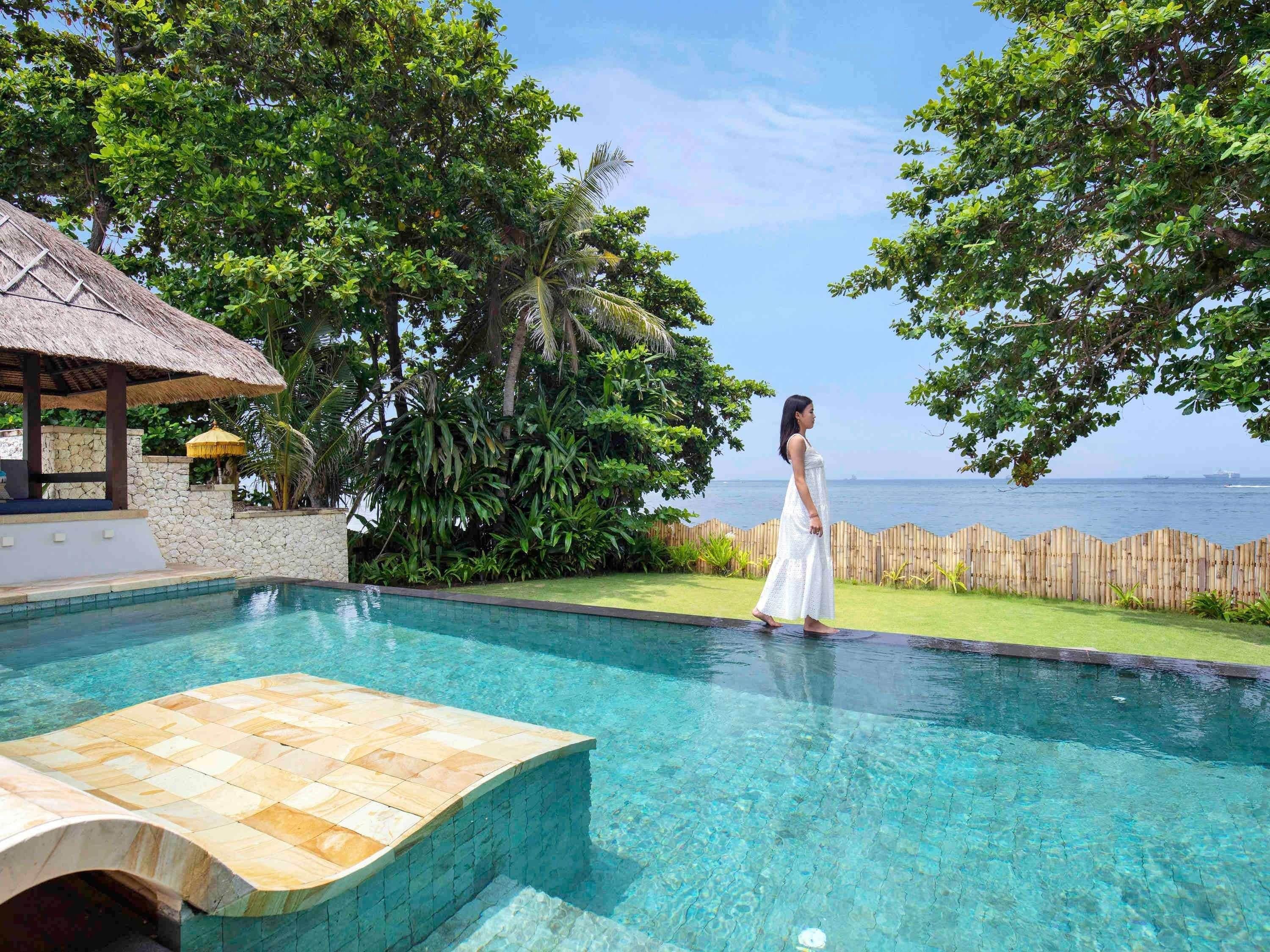 Uma área de piscina com vista desobstruída para o mar, rodeada por vegetação exuberante e uma estrutura com telhado de palha.