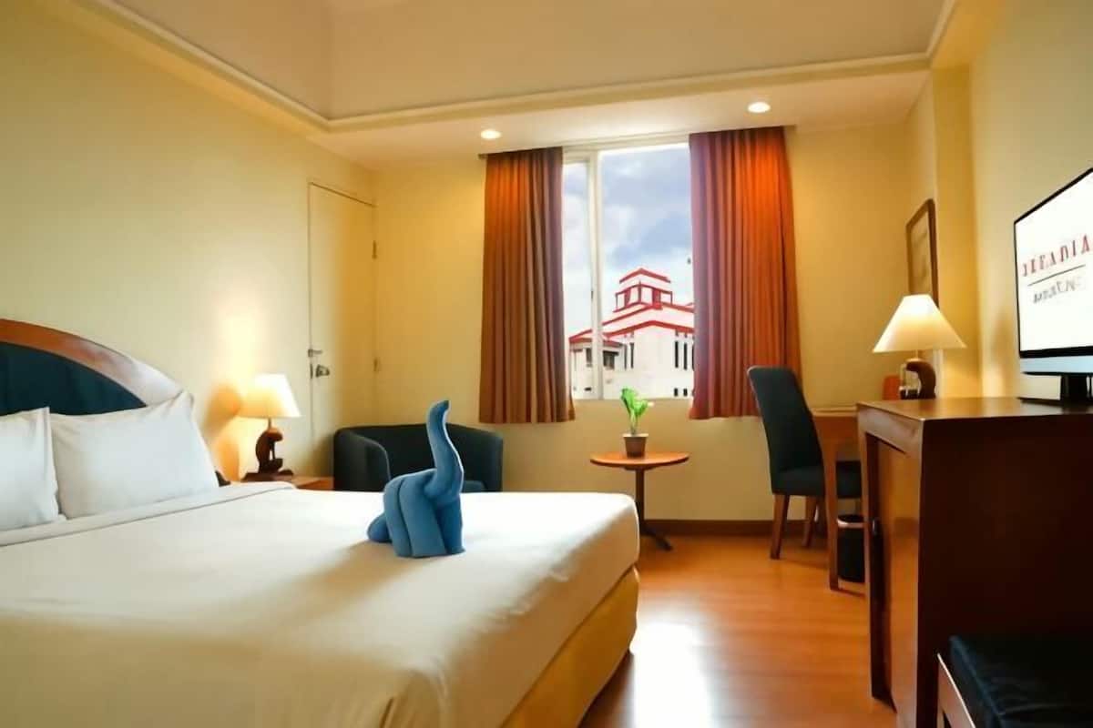 Quarto de hotel com cama, escrivaninha, cadeira, mesinha de apoio com abajur e vista para um edifício pela janela.