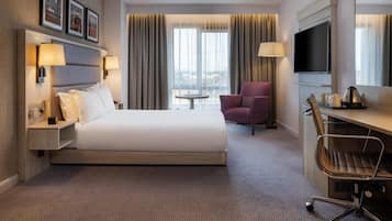 Quarto de hotel com cama, escrivaninha, cadeira, televisão e janela com cortinas.