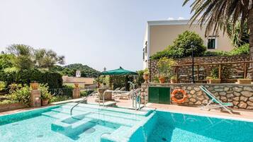 Piscina com degraus de acesso, rodeada por um muro de pedra e uma área de estar com cadeiras e guarda-sóis.