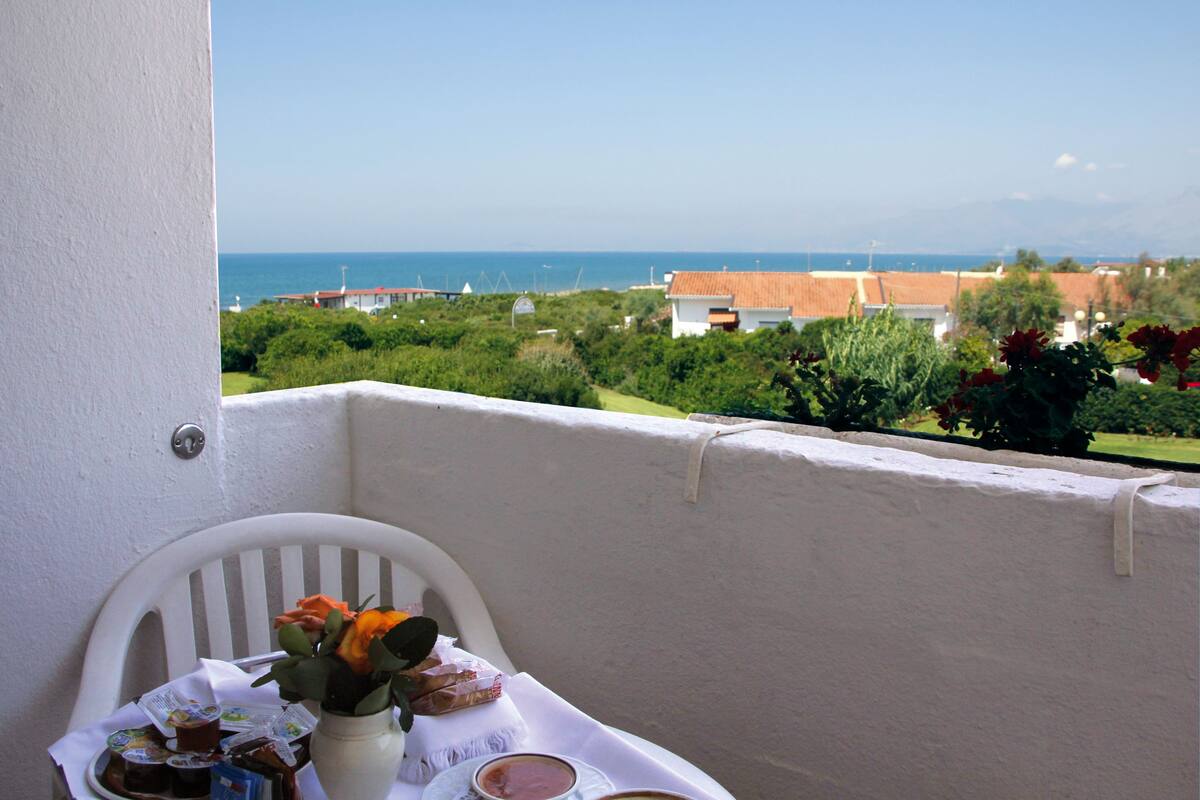 Varanda com mesa posta para dois, vista costeira com casas e vegetação.