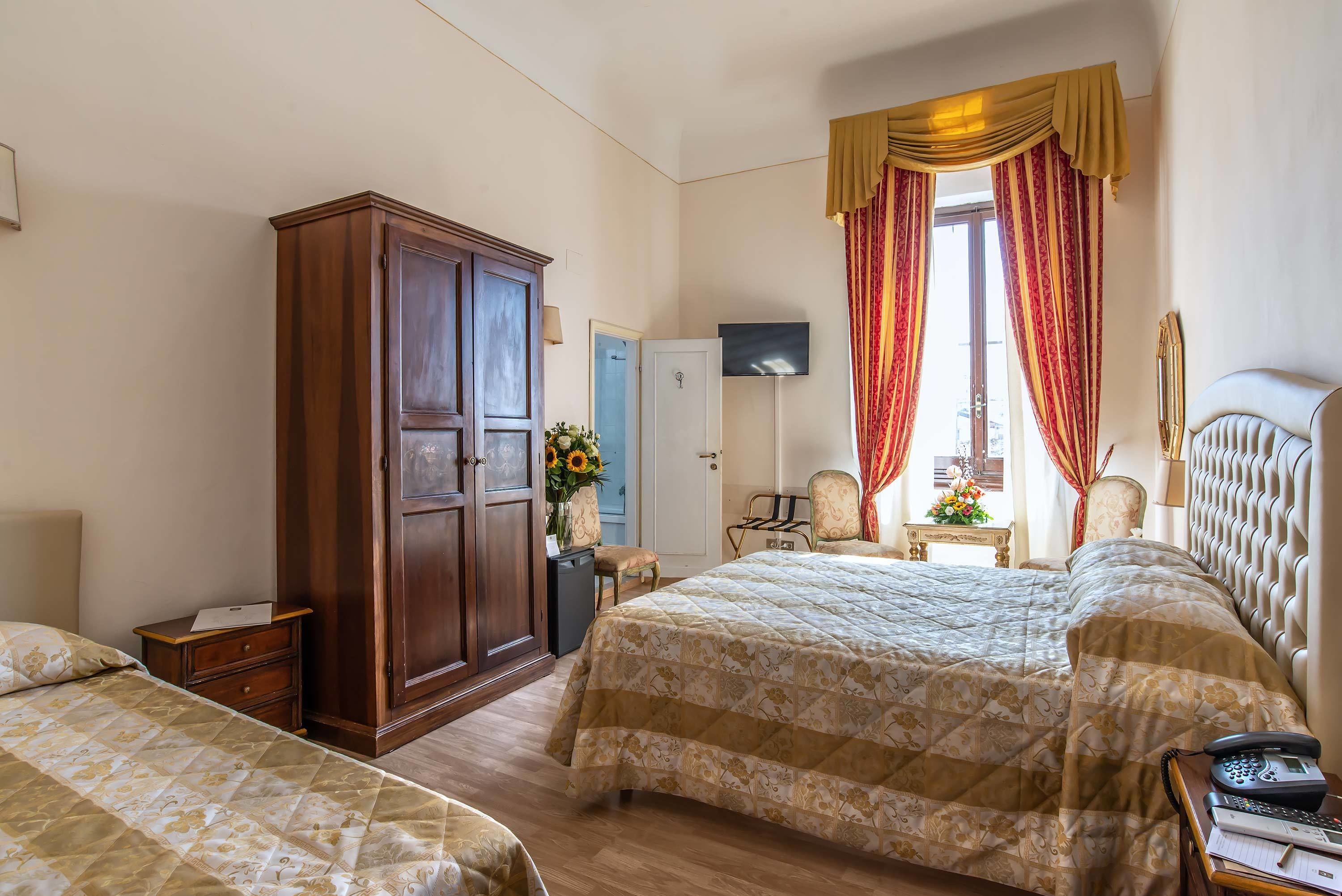 Quarto de hotel com duas camas, um guarda-roupa, uma televisão, uma mesa com telefone e uma janela com cortinas.
