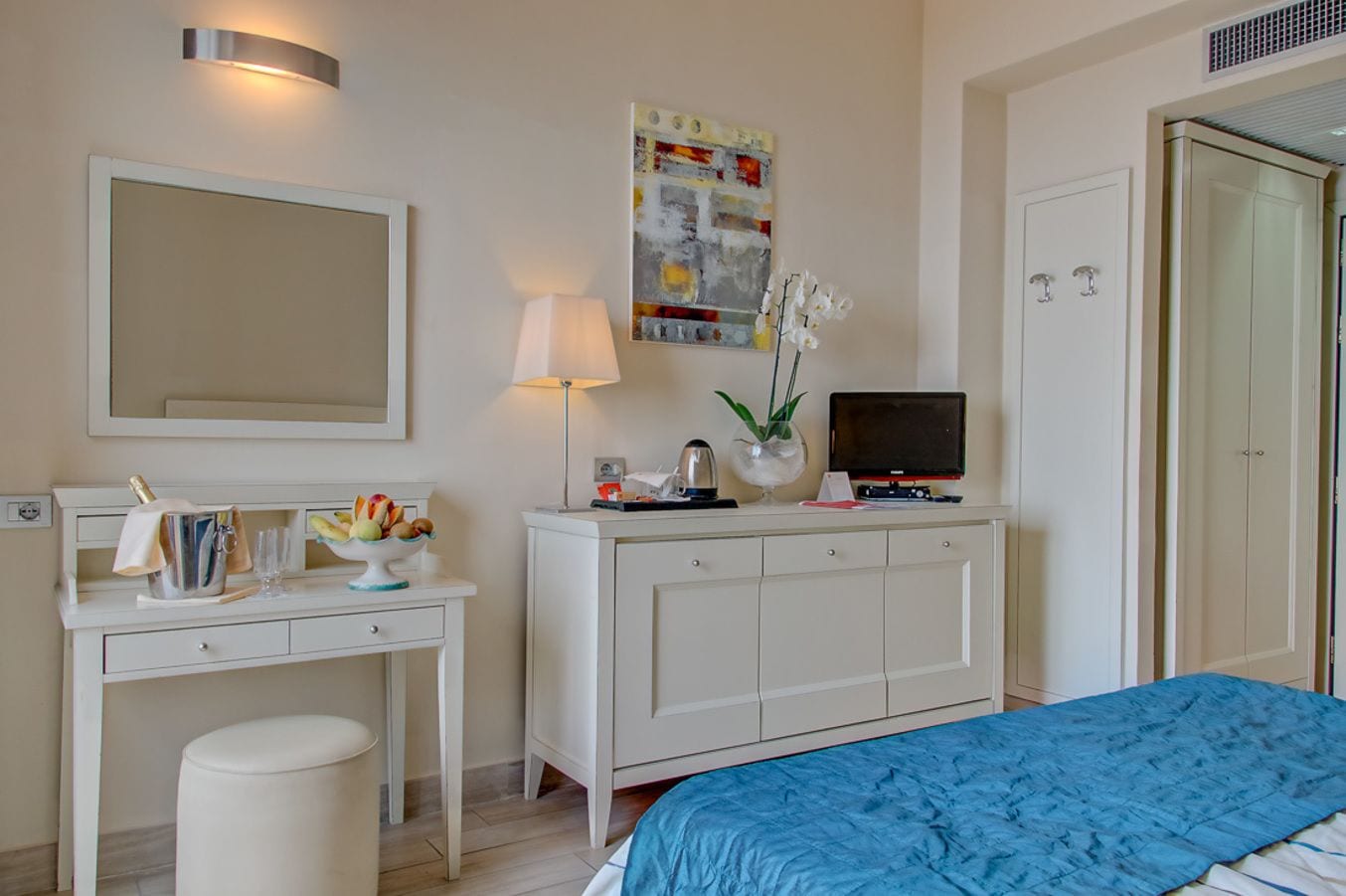 Quarto de hotel com cama, secretária com computador, espelho, luminária e um objeto decorativo na parede.