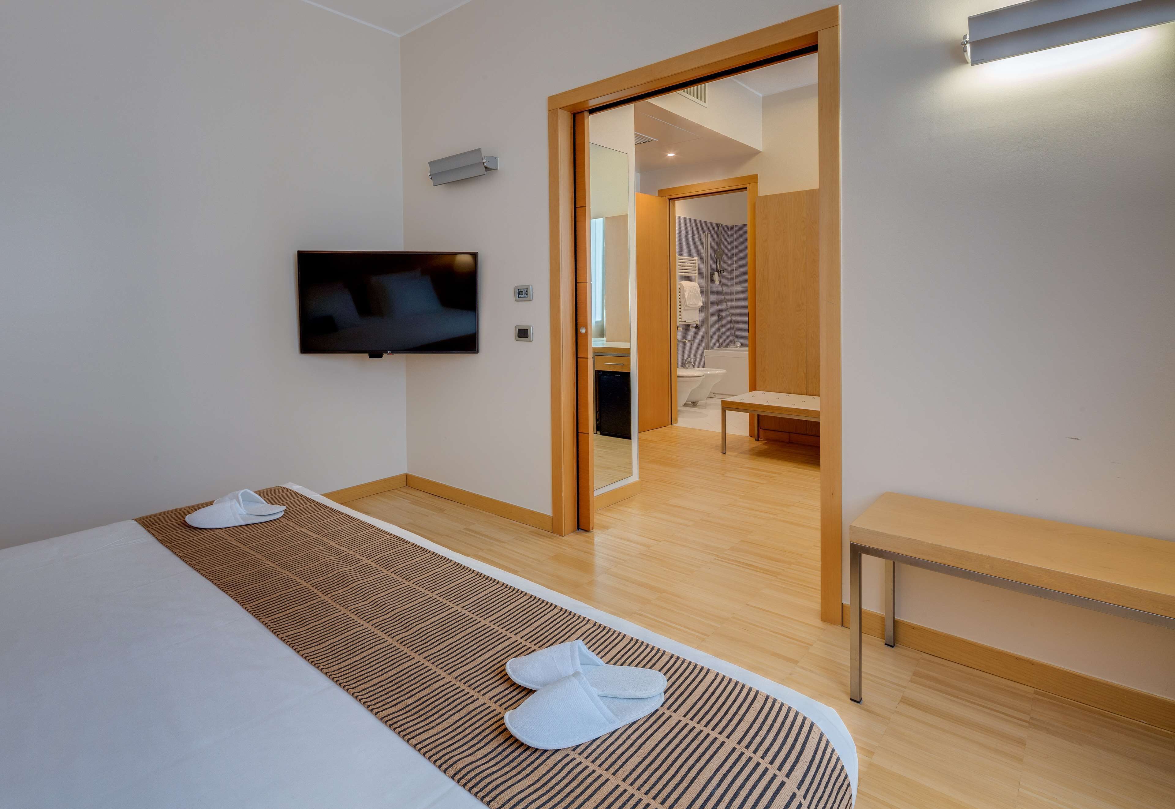 Quarto de hotel com cama, televisão, um banco de madeira e banheiro visível por uma porta aberta.