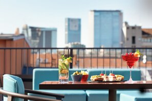 Um bar no terraço com vista para o horizonte da cidade, uma mesa com bebidas e petiscos, e cadeiras azuis.