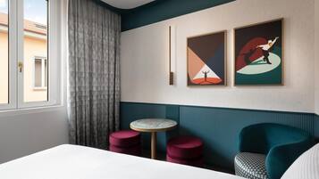 Quarto de hotel com cama, mesa redonda, dois banquinhos vermelhos, poltrona verde-água e duas pinturas abstratas na parede.