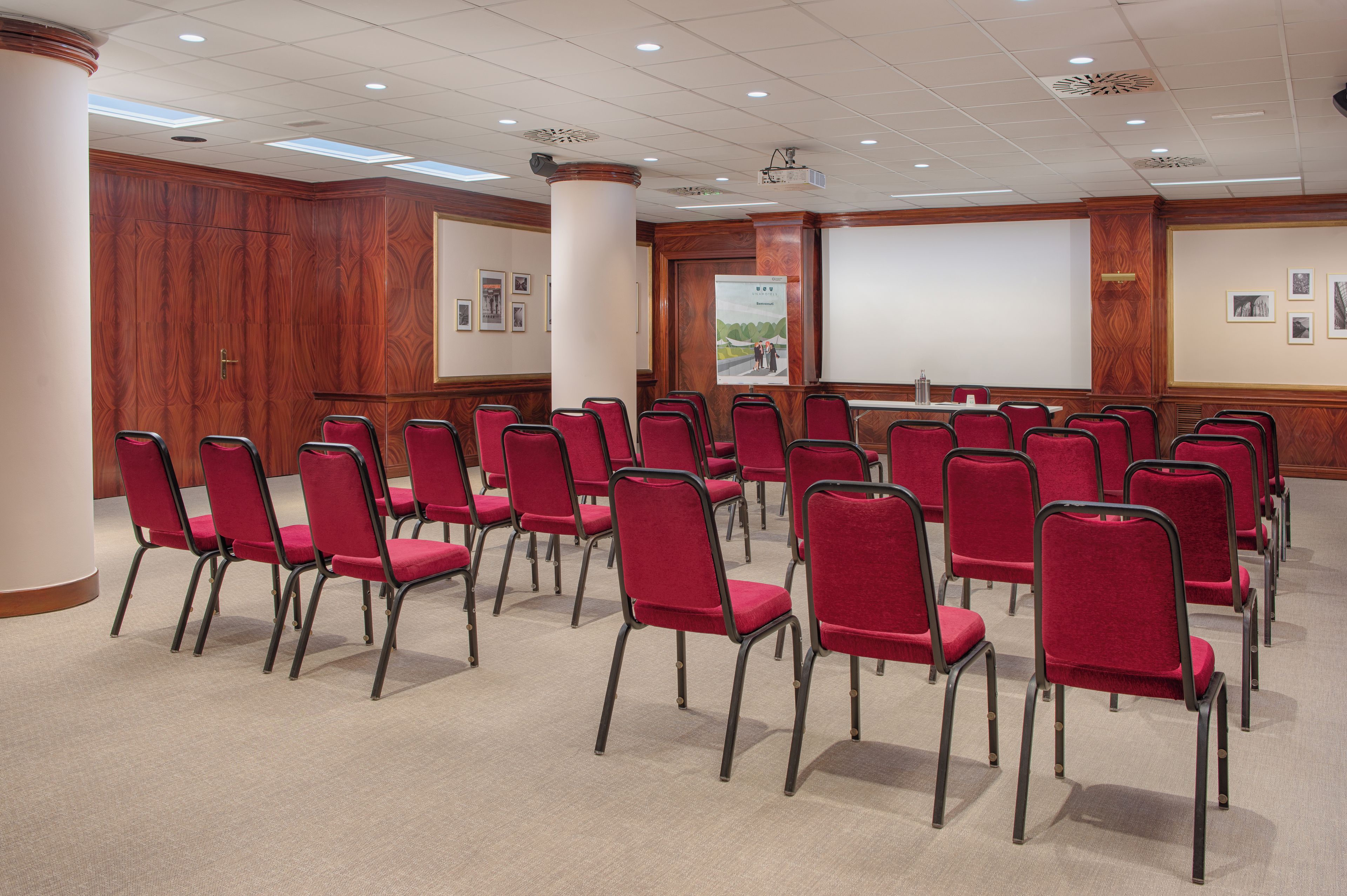 Uma sala de conferências com fileiras de cadeiras vermelhas, uma tela de projetor e uma parede revestida de madeira com quadros.