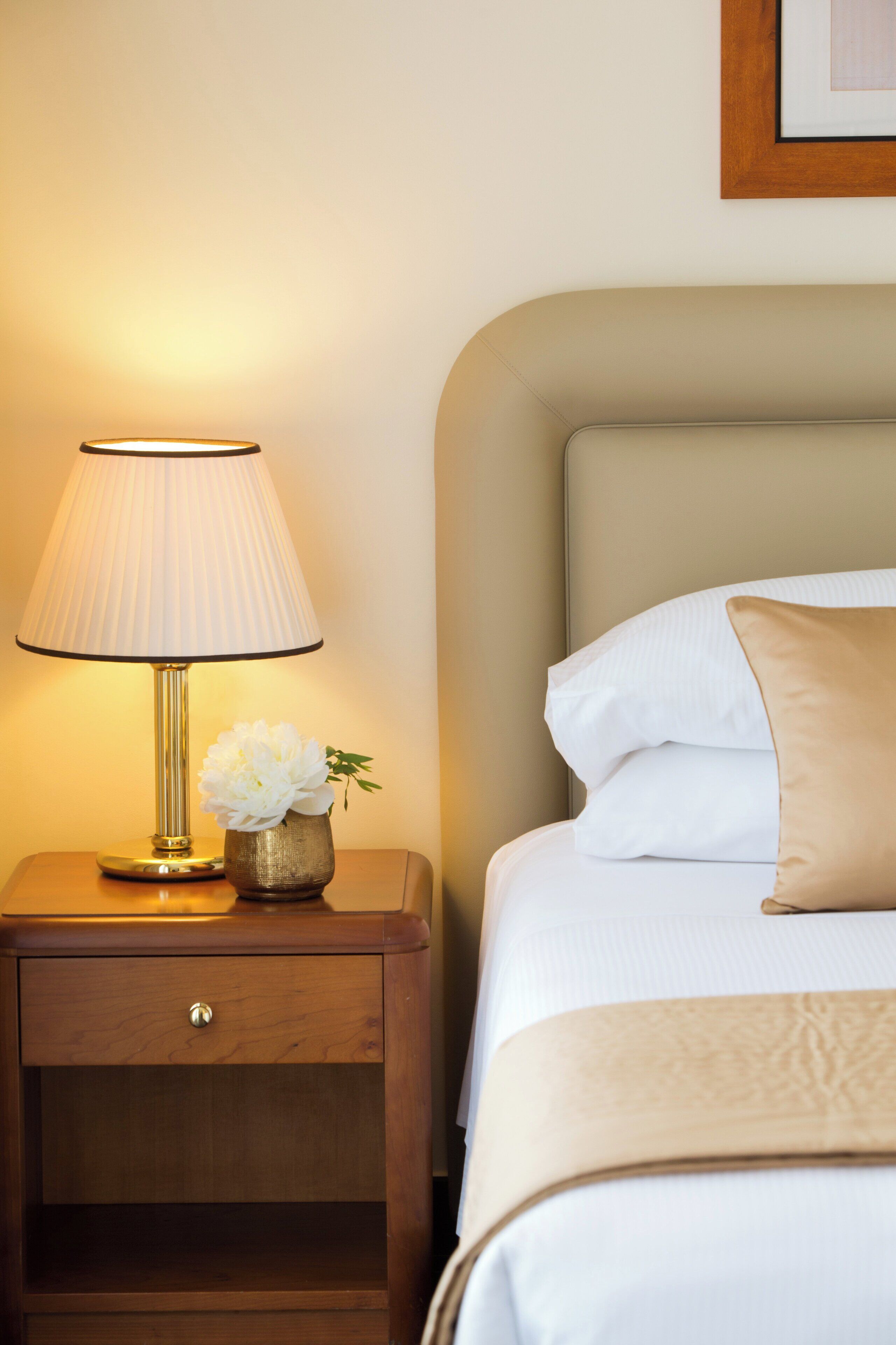 Cama bem arrumada com lençóis brancos e travesseiro bege, luminária de cabeceira e pequeno vaso com flores brancas em um criado-mudo de madeira.