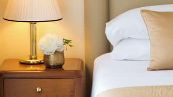 Cama bem arrumada com lençóis brancos e travesseiro bege, luminária de cabeceira e pequeno vaso com flores brancas em um criado-mudo de madeira.