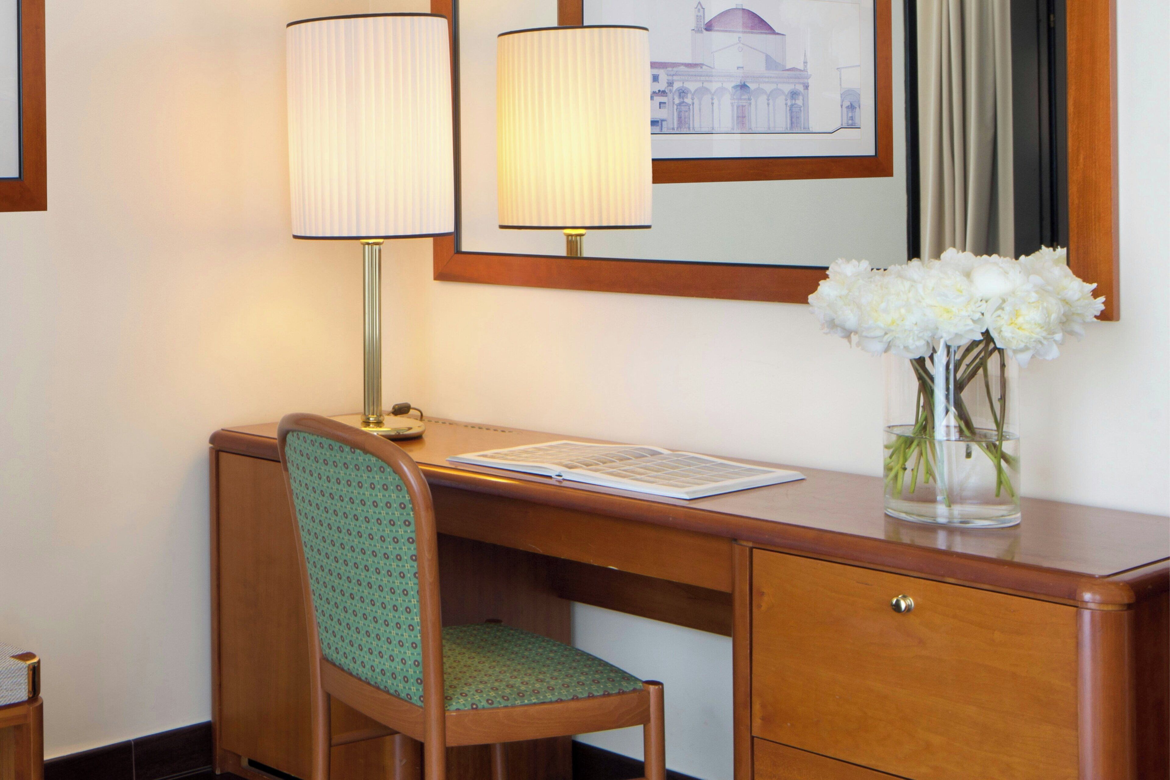 Mesa de hotel com uma cadeira, uma luminária, um vaso de flores e um quadro na parede.