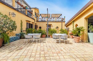 Um pátio com piso de azulejos, mobiliário de exterior, plantas em vasos e um edifício com varandas.