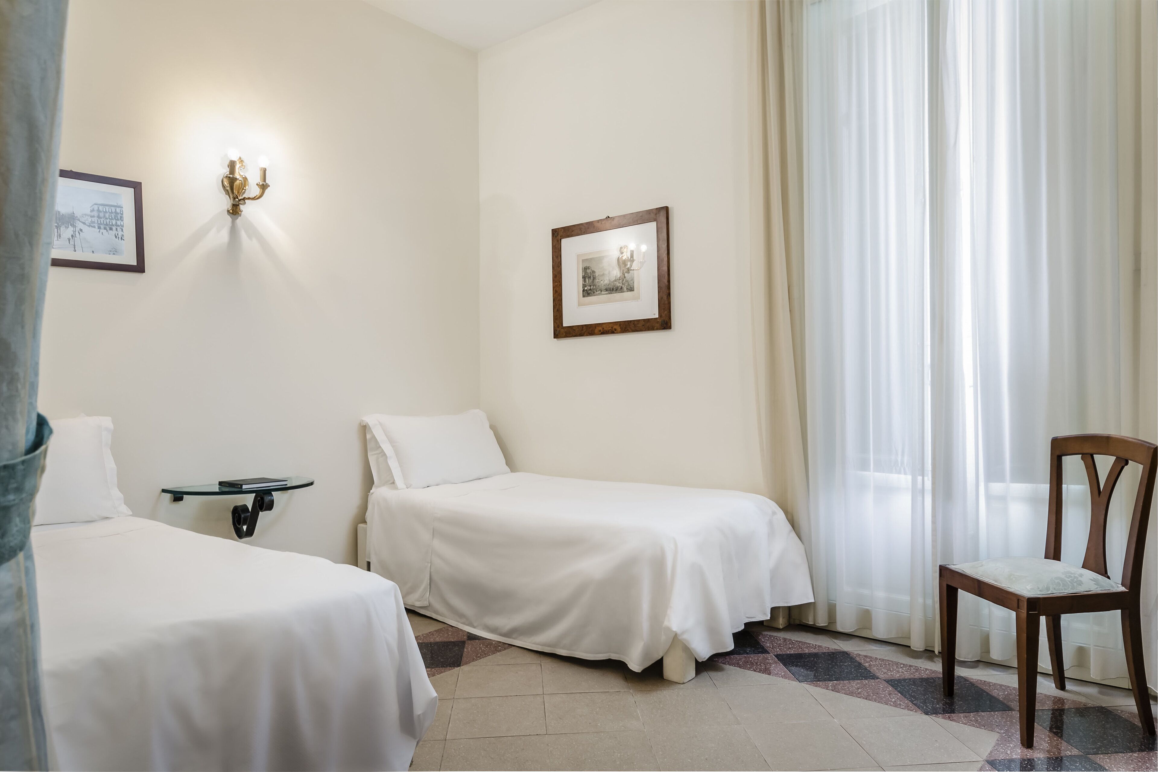 Quarto de hotel com duas camas de solteiro, uma mesinha, uma cadeira e um candeeiro fixado na parede.