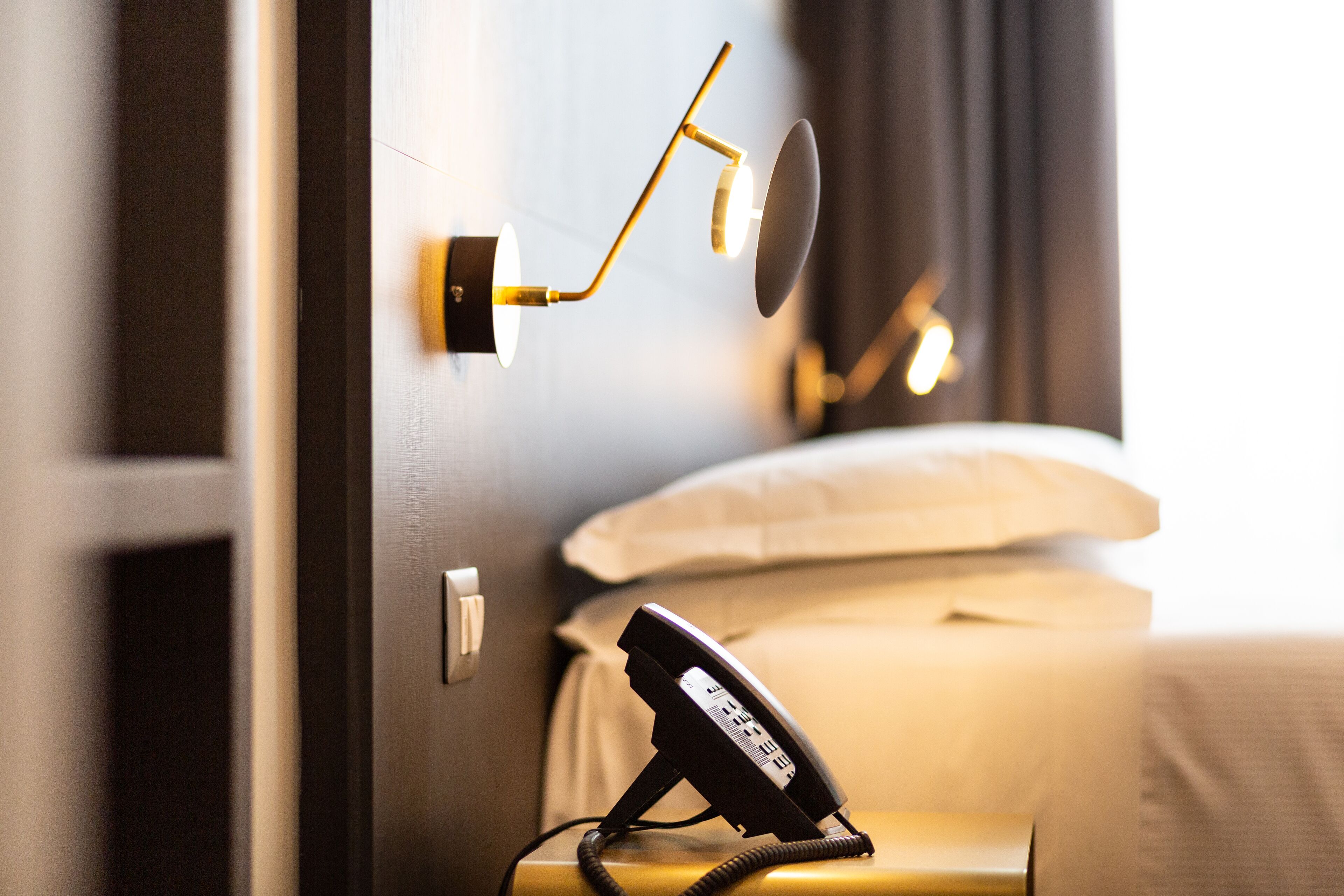 Quarto de hotel com telefone de mesa, luminária na parede e cama com travesseiro.