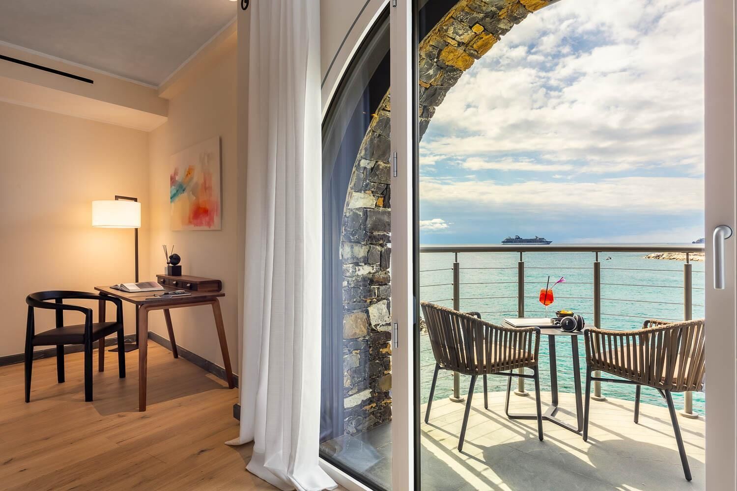 Quarto de hotel com secretária de madeira, cadeiras, uma luminária e uma grande janela com vista para o mar.