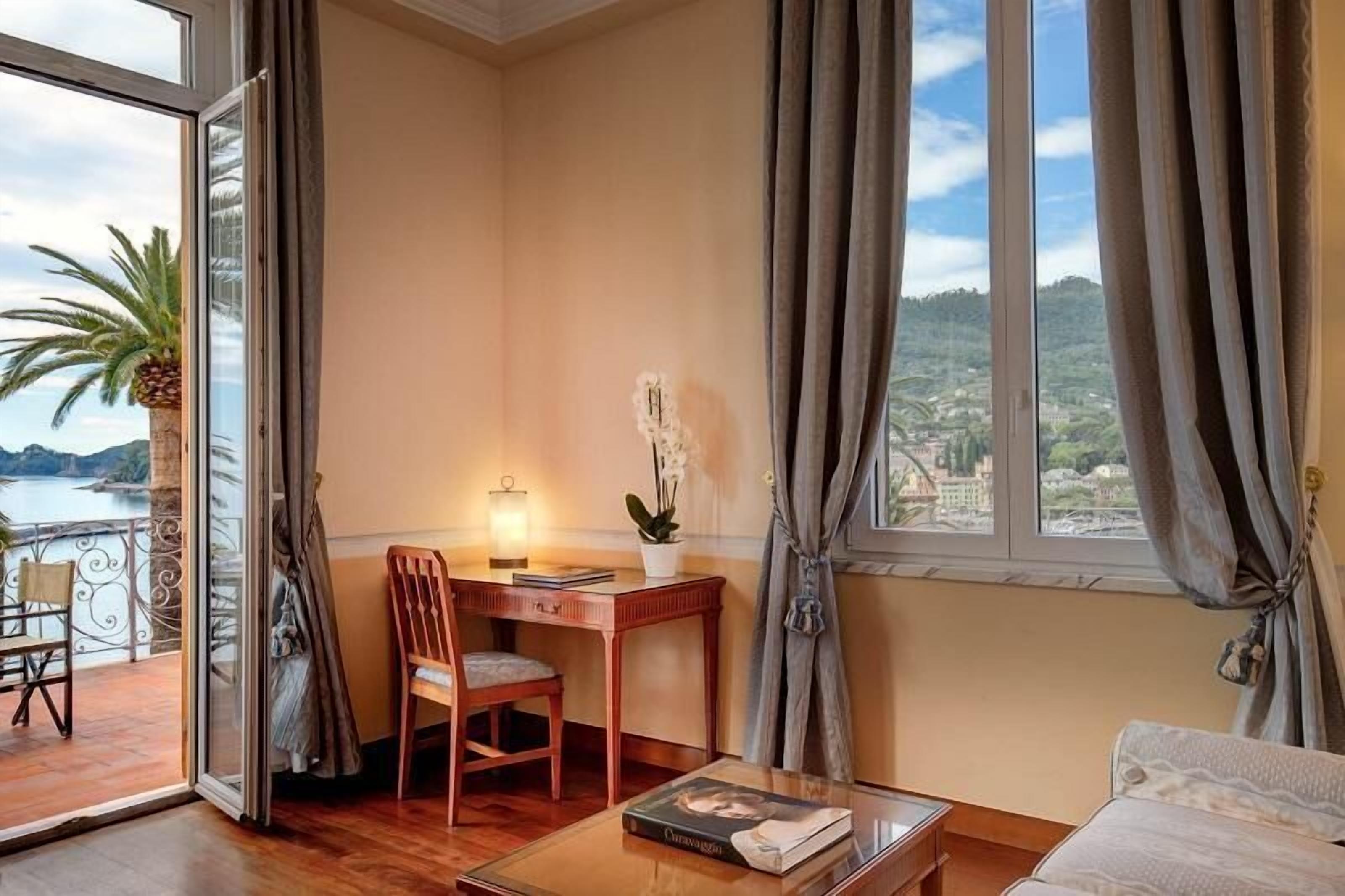 Quarto com escrivaninha de madeira, cadeira e vista para o mar através de uma grande janela.