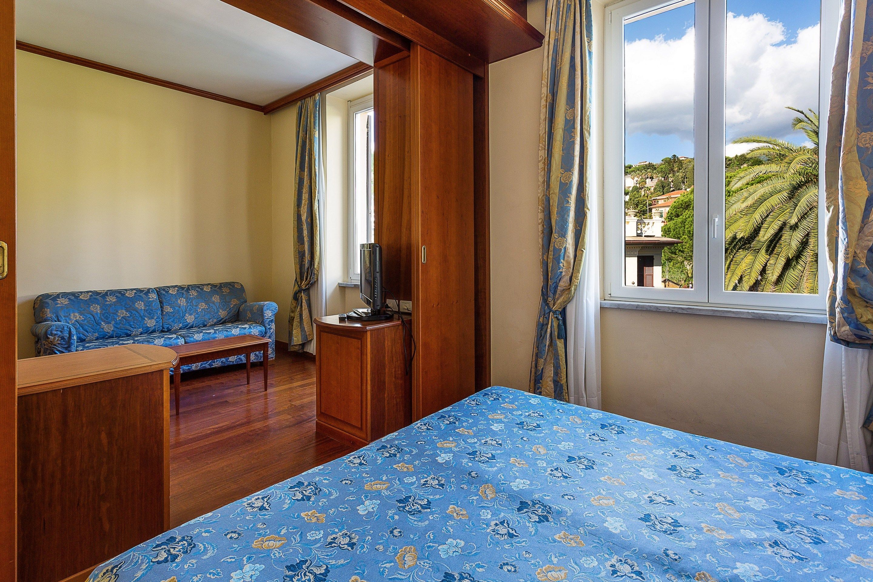Quarto com cama, sofá, televisão e janela com vista para uma colina.