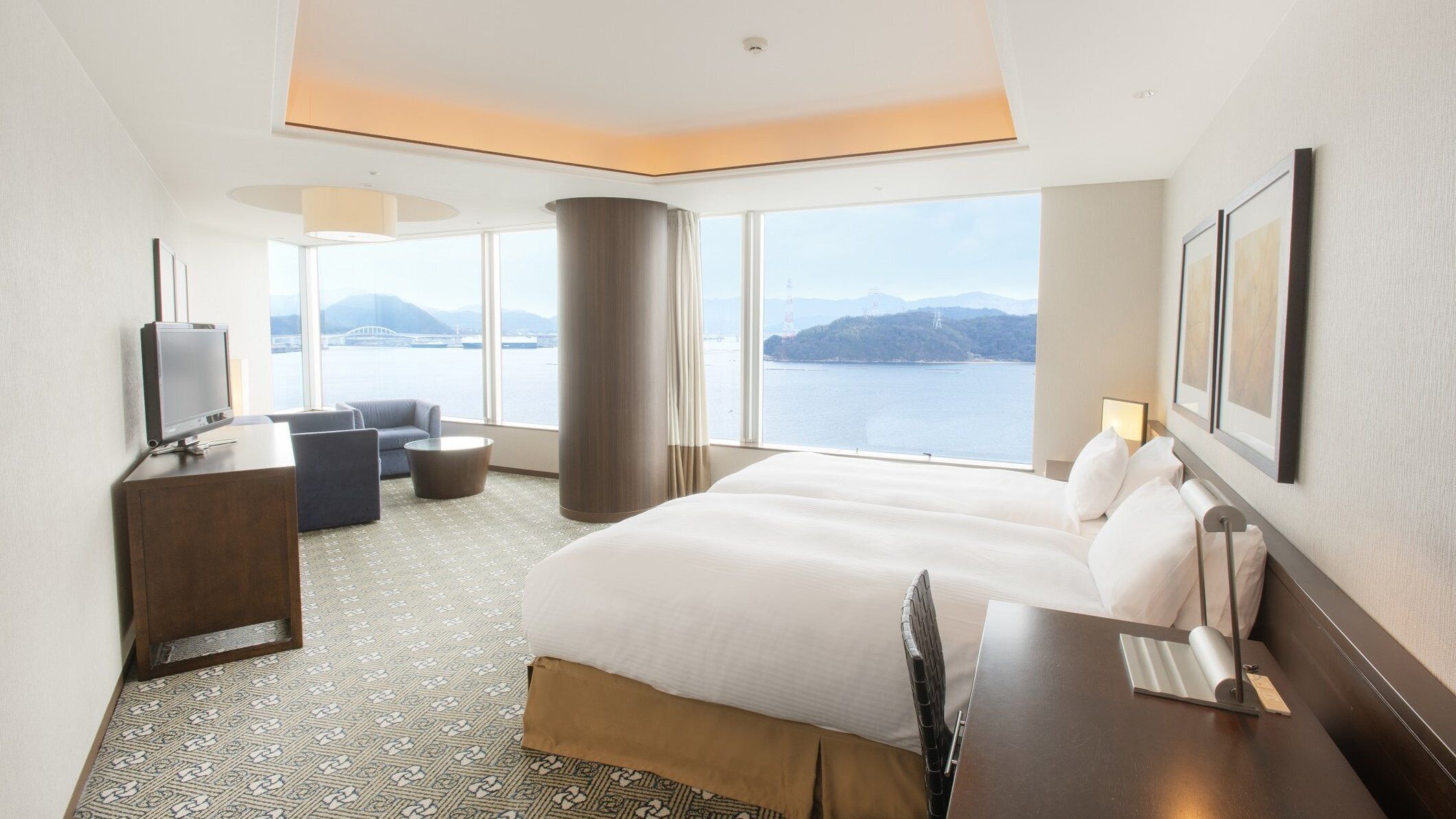 Quarto de hotel com uma cama grande, uma mesa de trabalho com televisão, uma cadeira, uma mesinha e vista para um corpo de água e montanhas.