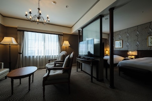 Quarto de hotel com uma cama grande, televisão, escrivaninha com cadeira, sofá e lustre.