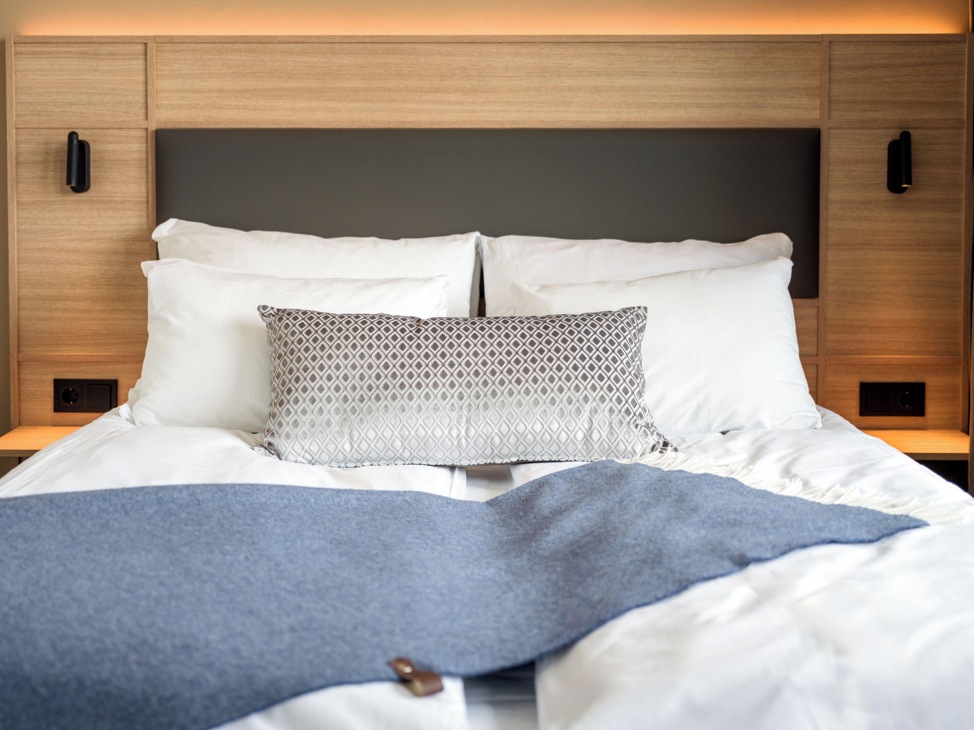 Cama bem arrumada com lençóis brancos, almofada cinza estampada e manta azul.