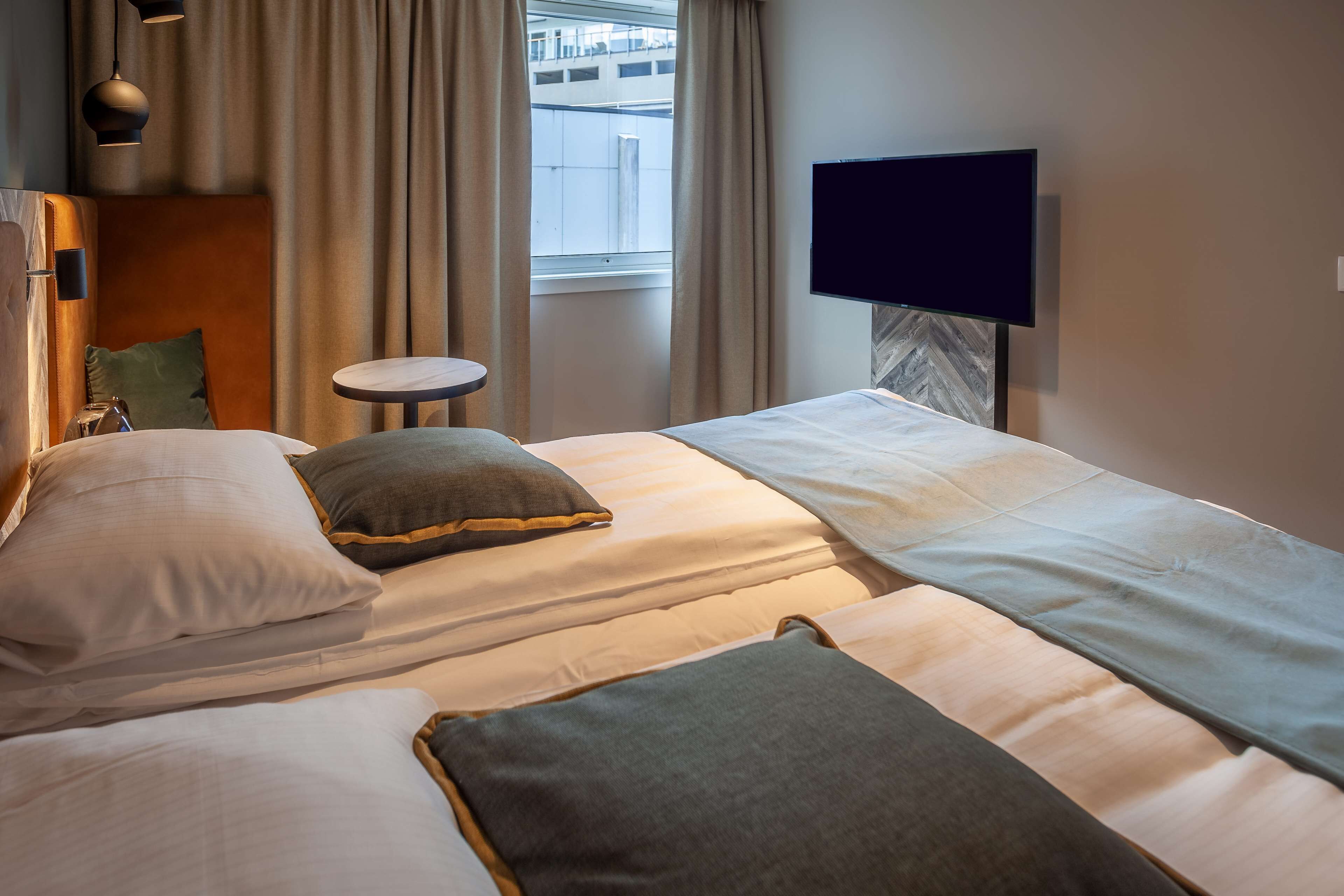 Quarto de hotel com duas camas, uma mesa redonda pequena, televisão fixada na parede e uma janela com cortinas.