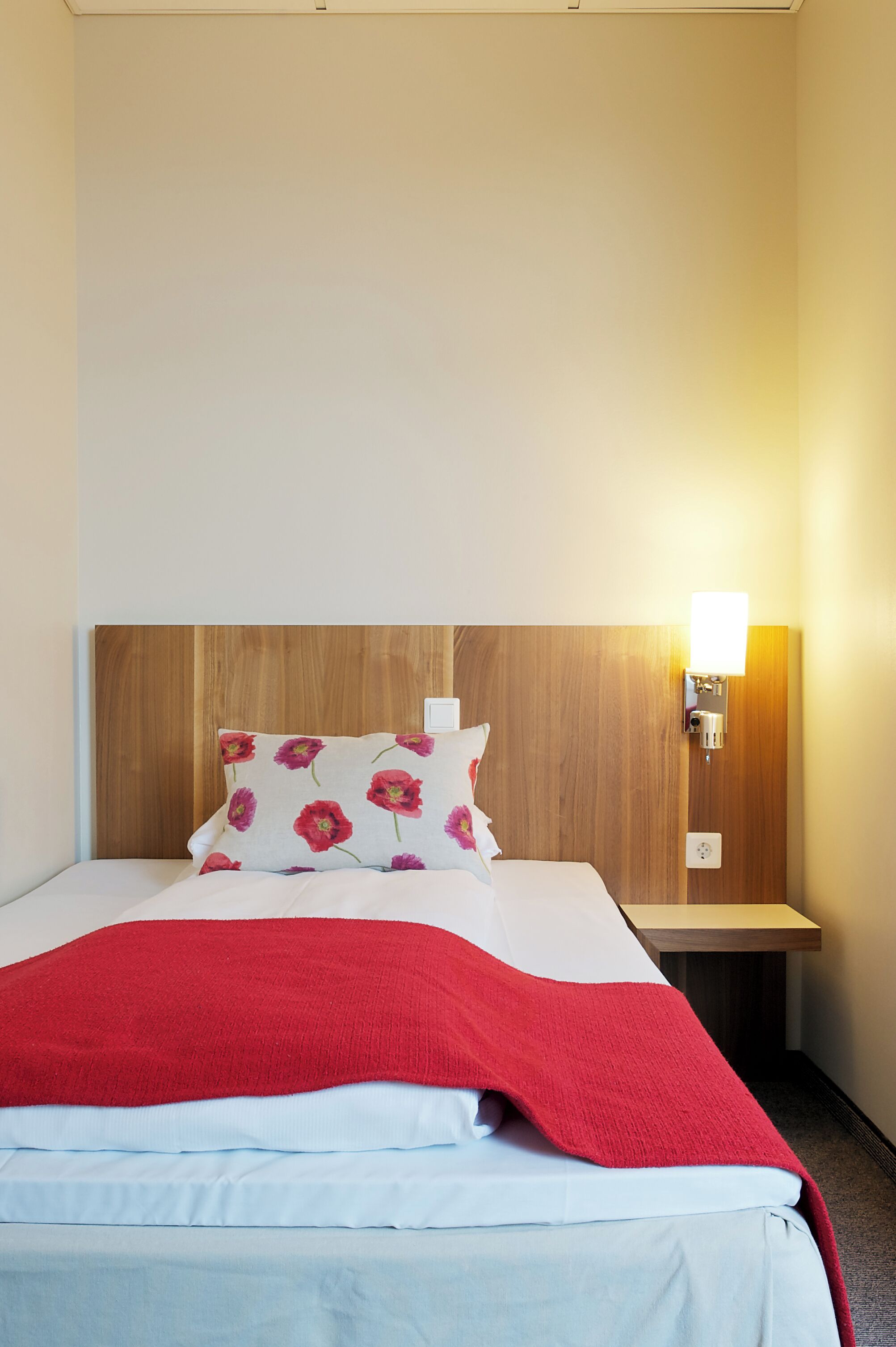 Quarto de hotel com cabeceira de madeira, manta vermelha e luminária fixada na parede.