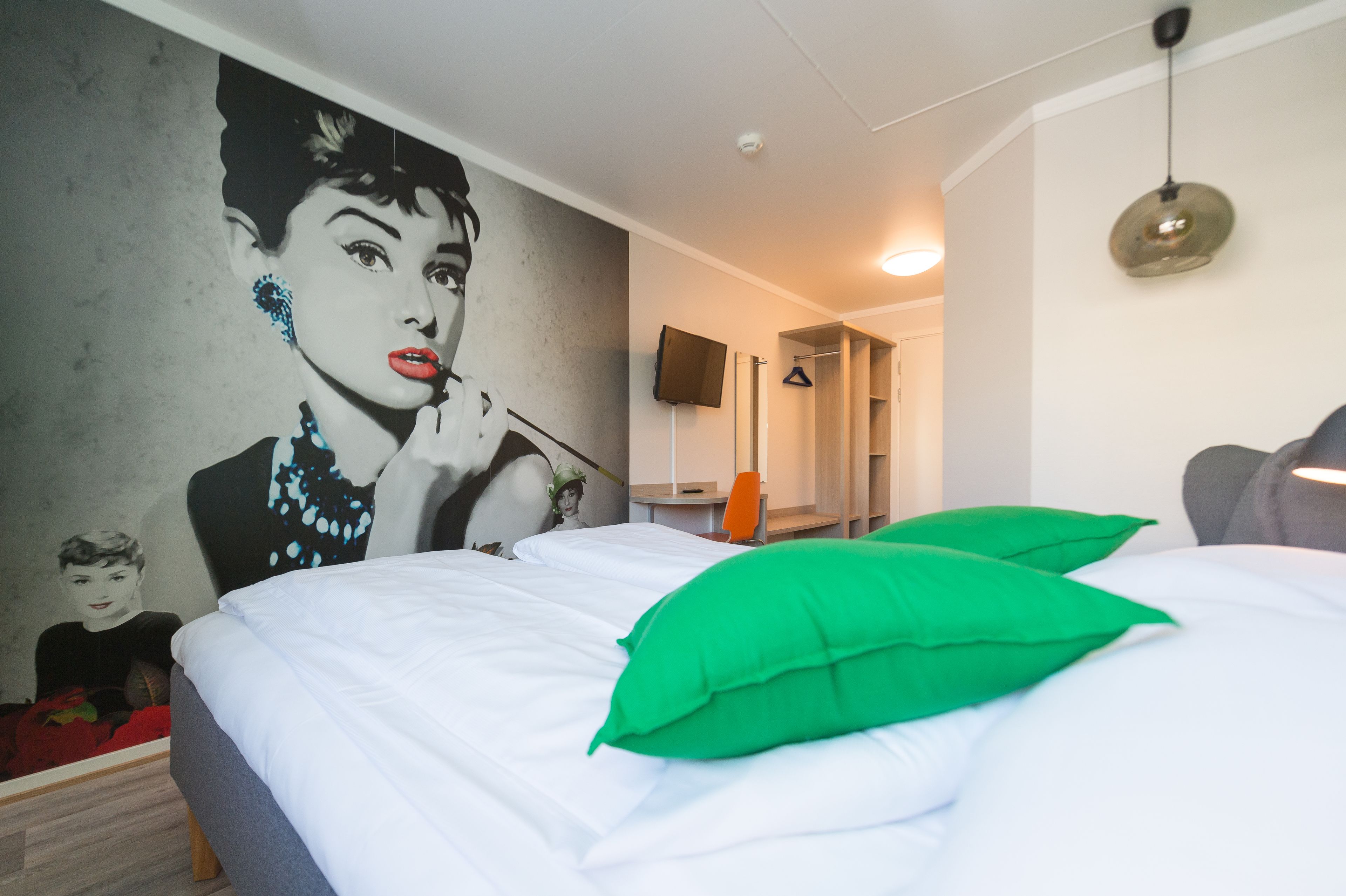 Quarto de hotel moderno com uma cama grande, almofadas verdes, televisão de tela plana e um mural na parede.