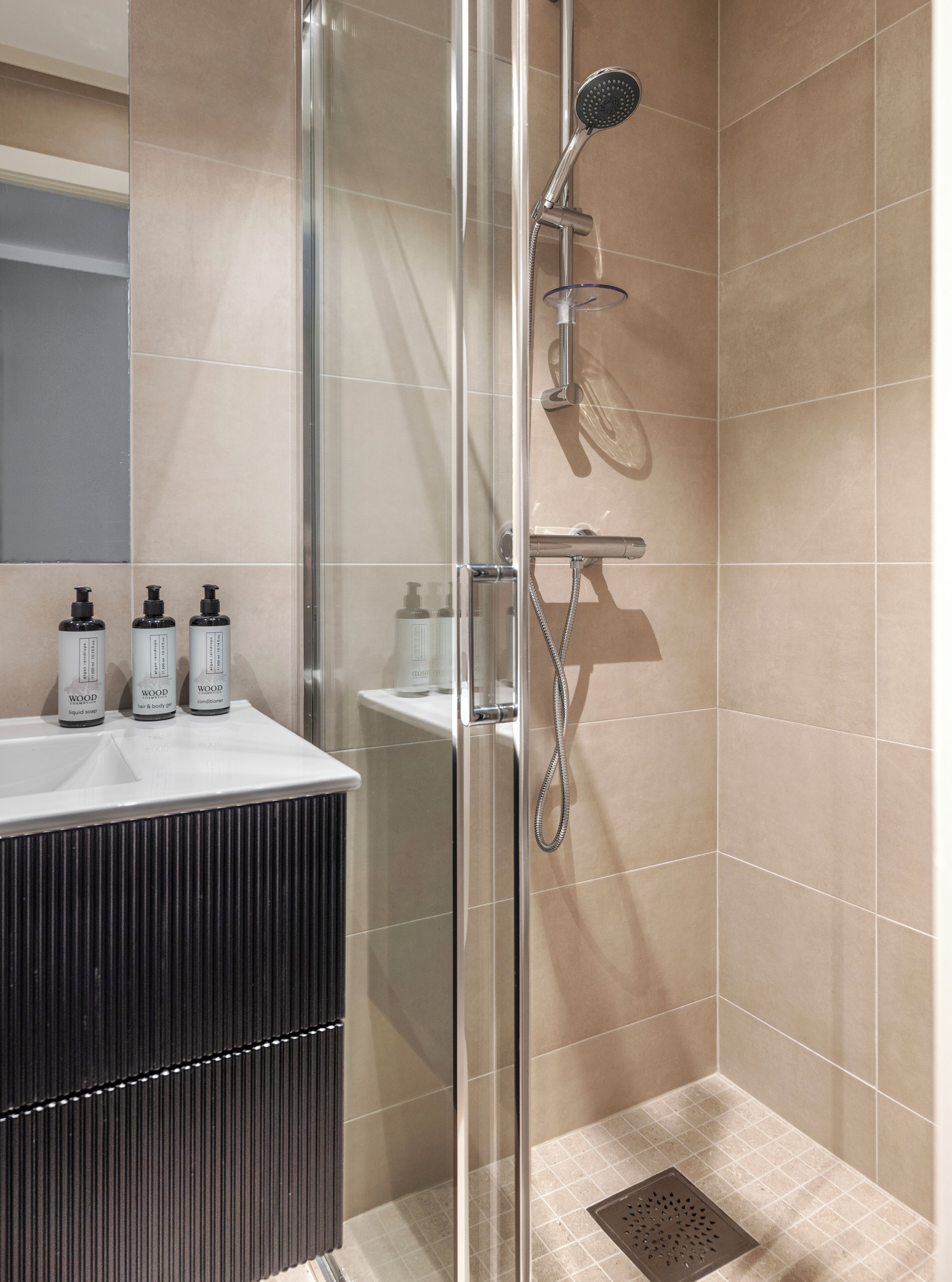 Um banheiro moderno com chuveiro envidraçado, um lavatório branco com três produtos da marca Wood e paredes revestidas de azulejos bege.