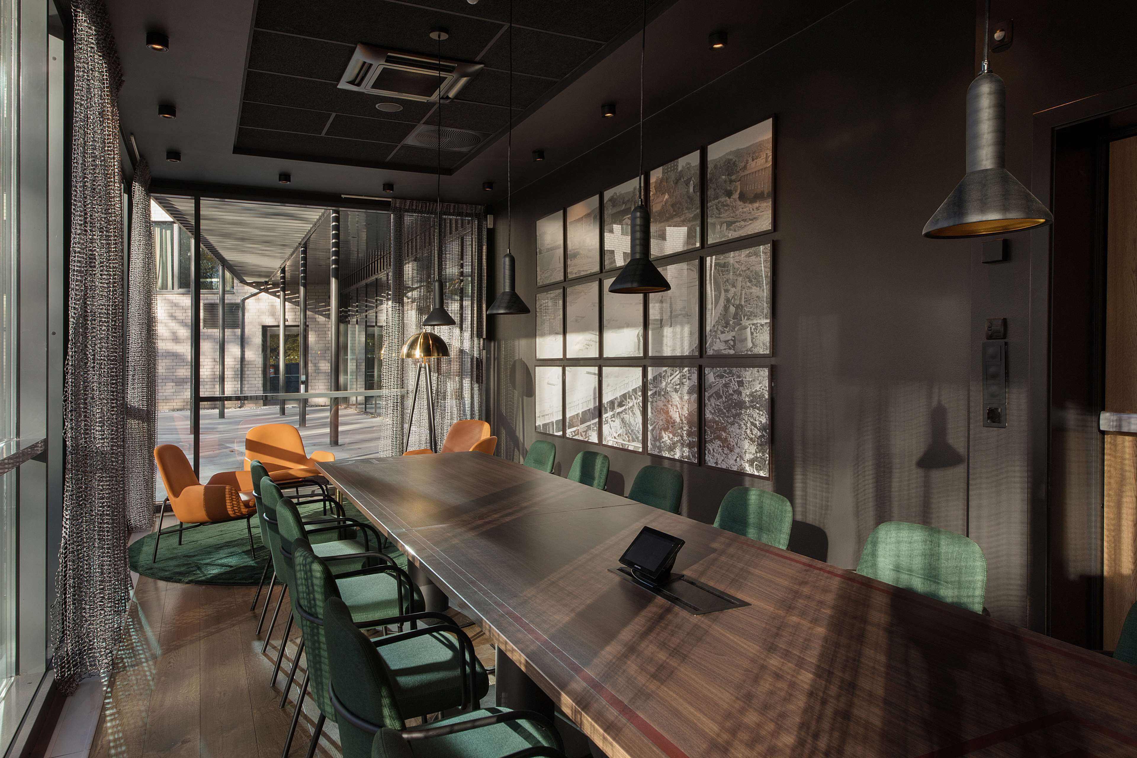 Sala de conferência com uma mesa comprida, cadeiras verdes e laranjas e luminárias pendentes.