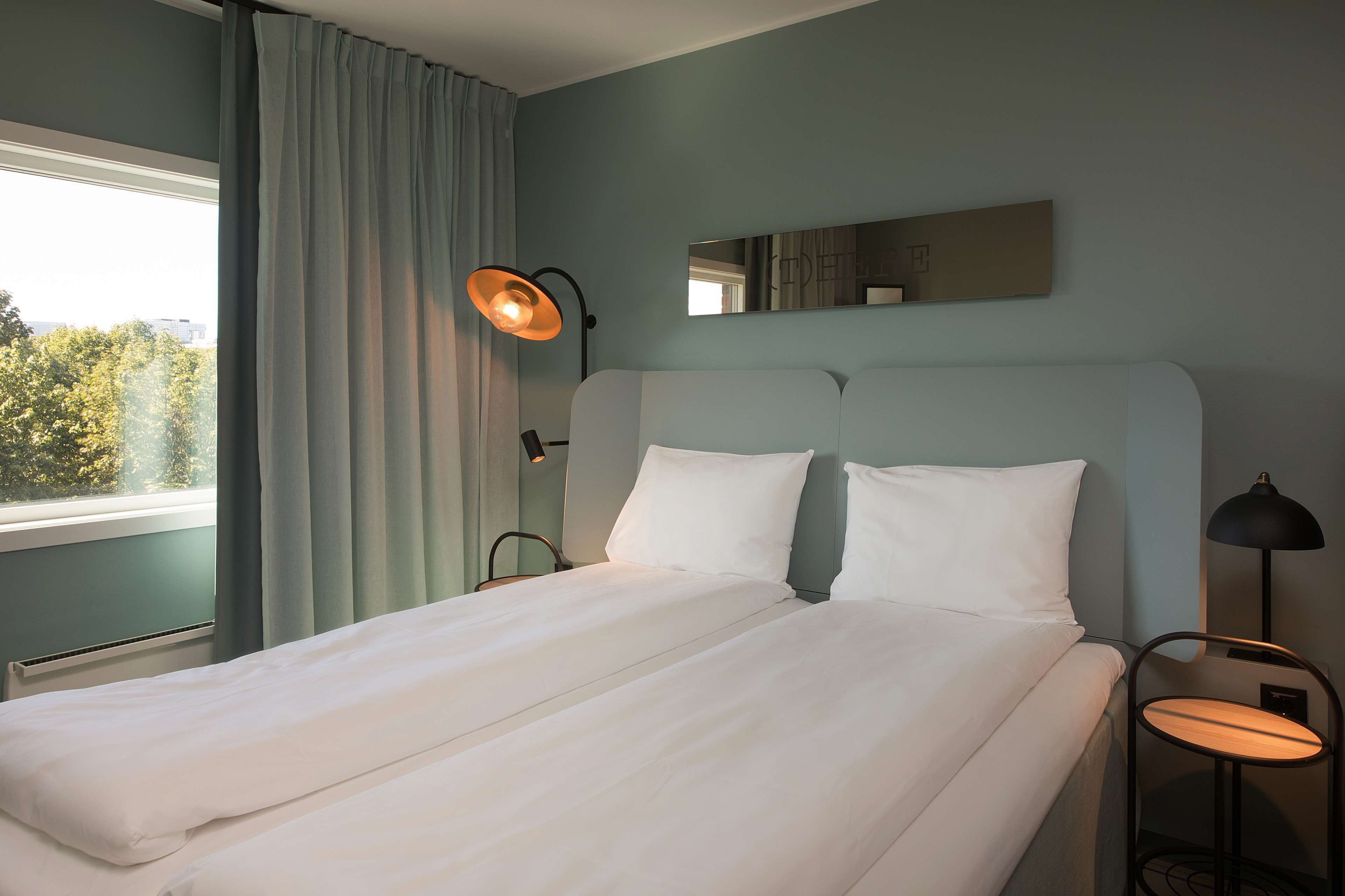 Cama bem arrumada com lençóis brancos, abajur e janela com cortinas.