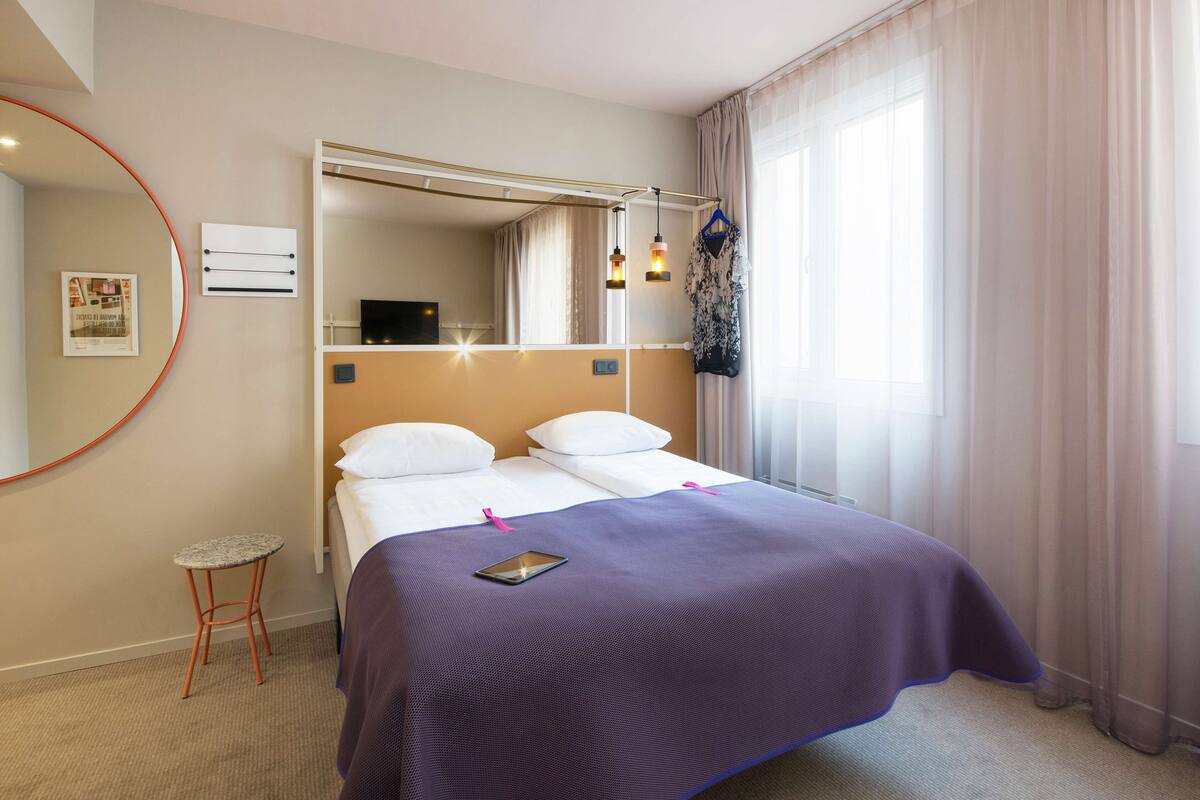 Quarto de hotel com cama, um banquinho, espelho e janela com cortinas.