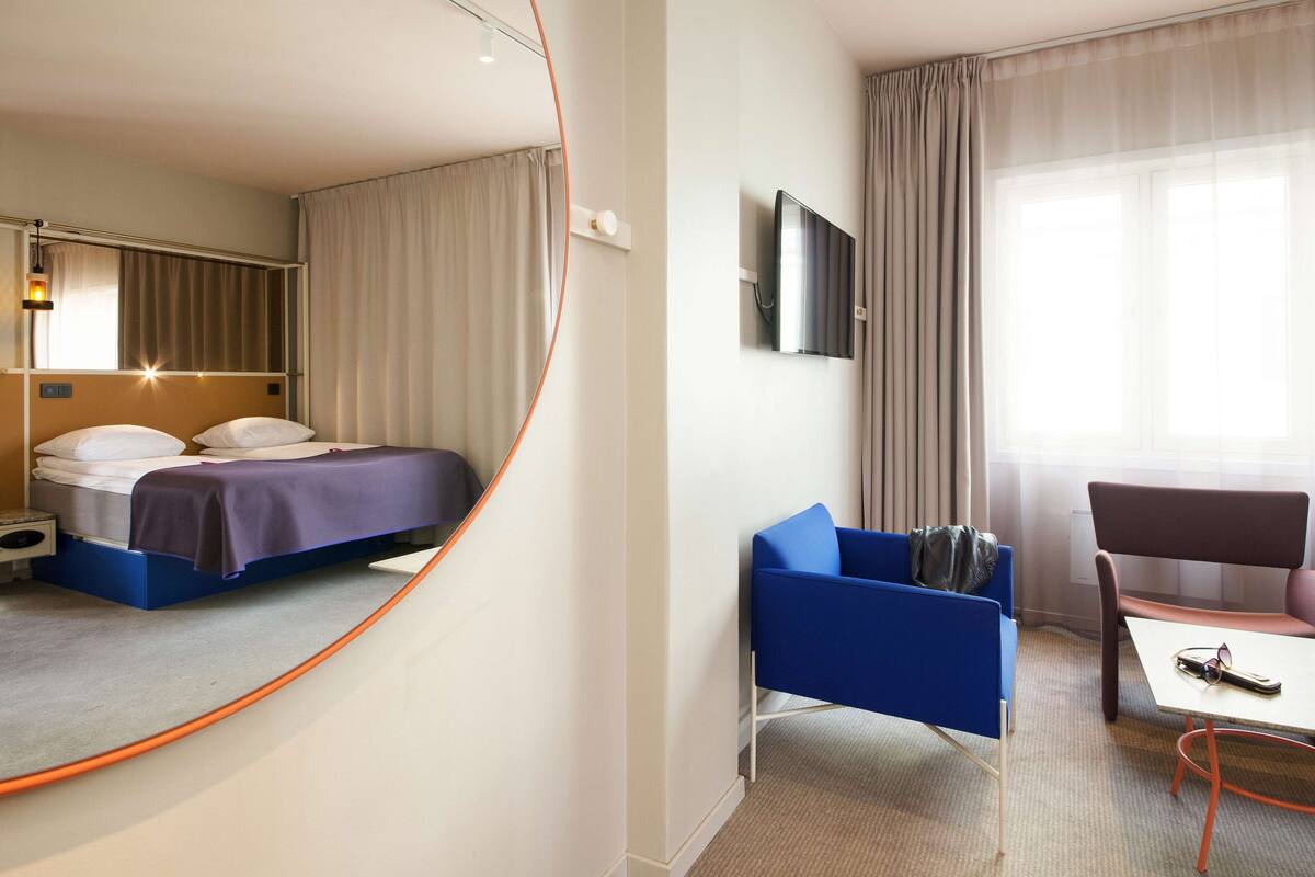 Quarto de hotel com cama, escrivaninha, cadeira, televisão e janela com cortinas.
