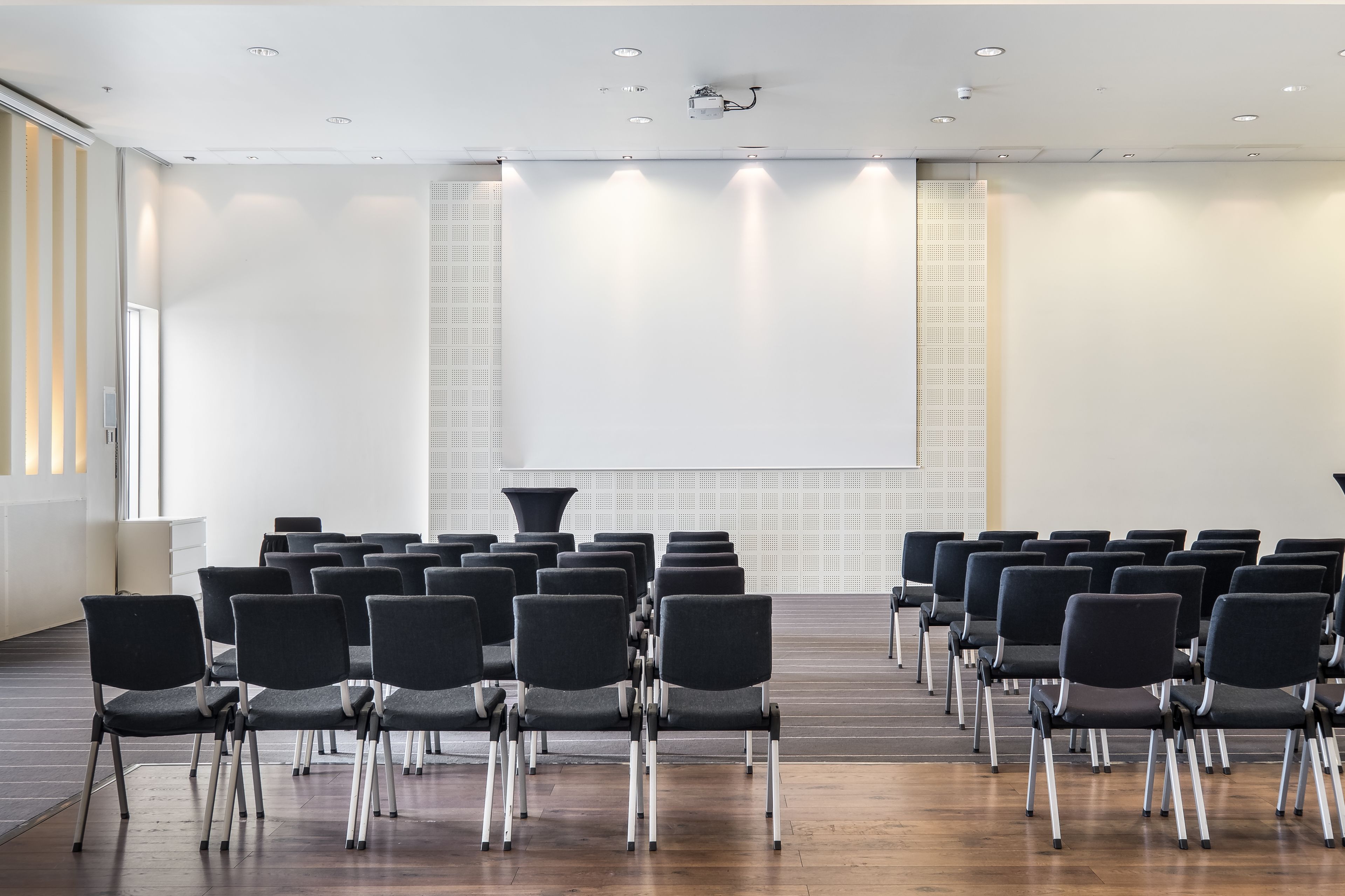 Uma sala de conferências com fileiras de cadeiras pretas, uma tela grande e um palanque.