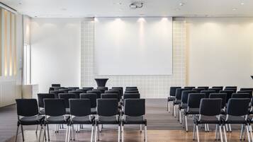 Uma sala de conferências com fileiras de cadeiras pretas, uma tela grande e um palanque.