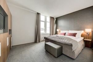 Quarto de hotel moderno com uma cama grande, uma mesa de cabeceira com abajur, uma TV de tela plana e uma janela com cortinas.