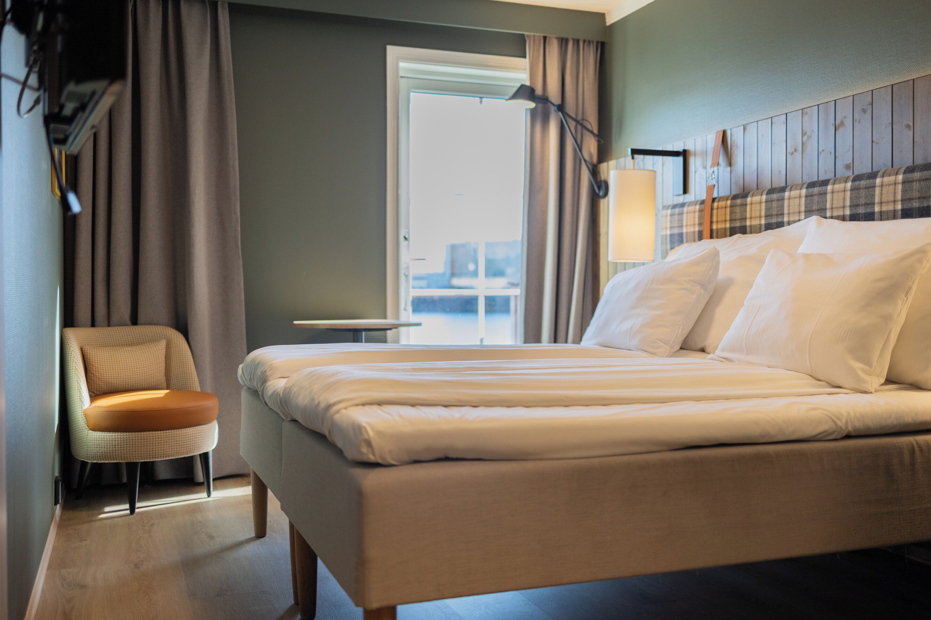 Quarto de hotel com cama, uma cadeira, luminária e vista para o exterior.