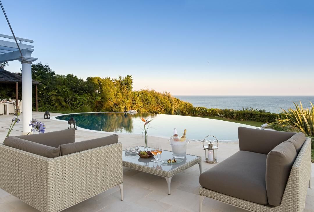 Uma área à beira da piscina com mobiliário de vime, uma mesa de vidro e vista para o mar.
