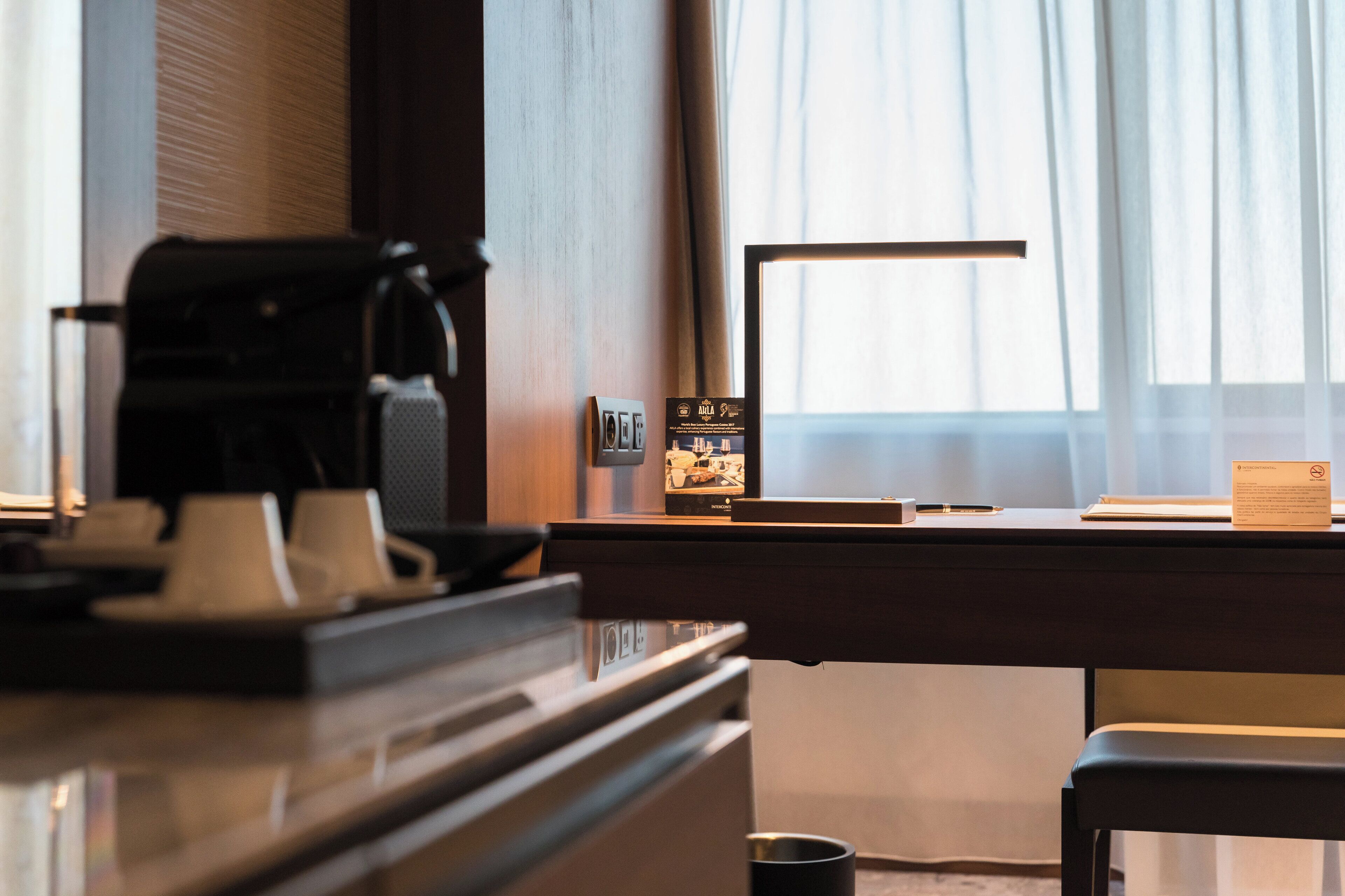 Quarto de hotel com escrivaninha, cafeteira, luminária e janela com cortinas.