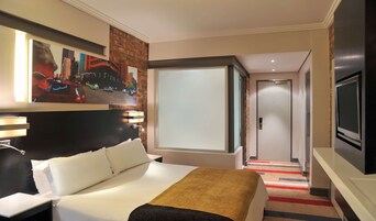 Quarto de hotel com uma cama, televisão, quadro e vista para a paisagem urbana.