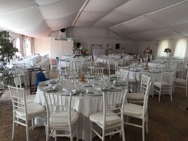 Espaço para eventos com tendas, mesas redondas preparadas para refeições, cadeiras brancas e centros de mesa florais.