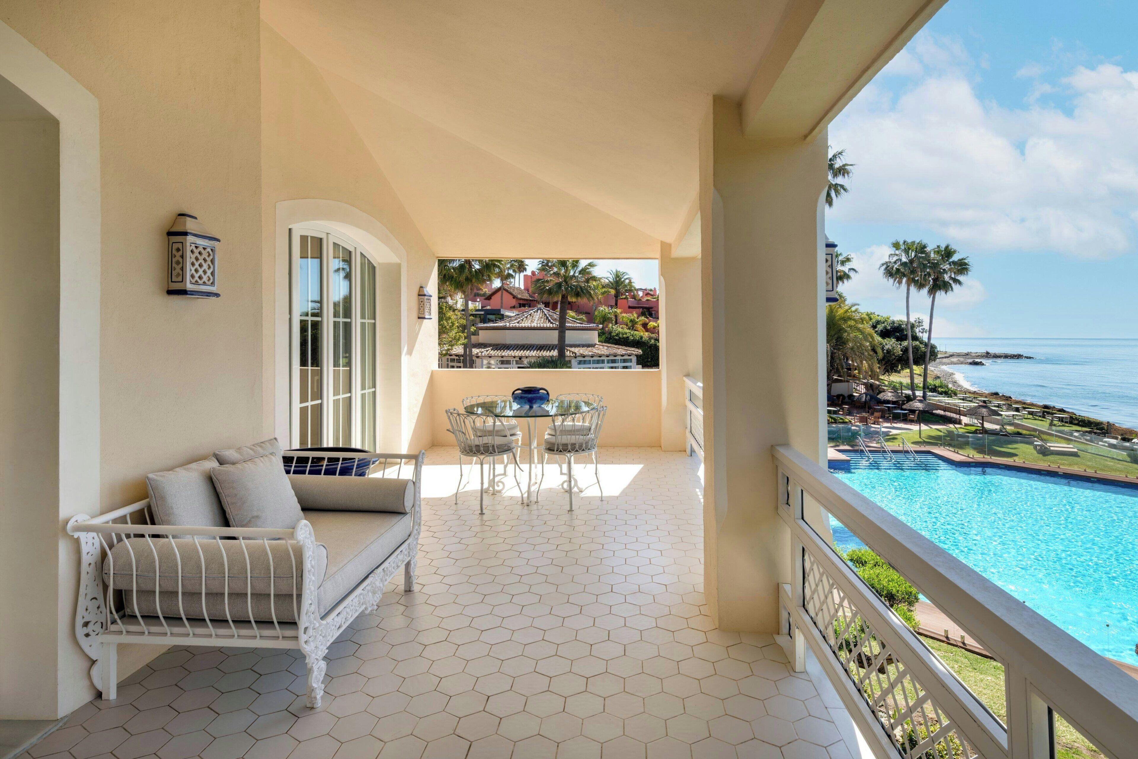 Varanda com sofá branco, mesa de vidro e cadeiras, com vista para uma piscina e o litoral.