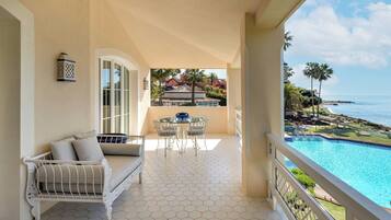 Varanda com sofá branco, mesa de vidro e cadeiras, com vista para uma piscina e o litoral.