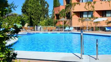 Piscina com espreguiçadeiras e guarda-sóis, cercada por uma grade e árvores.