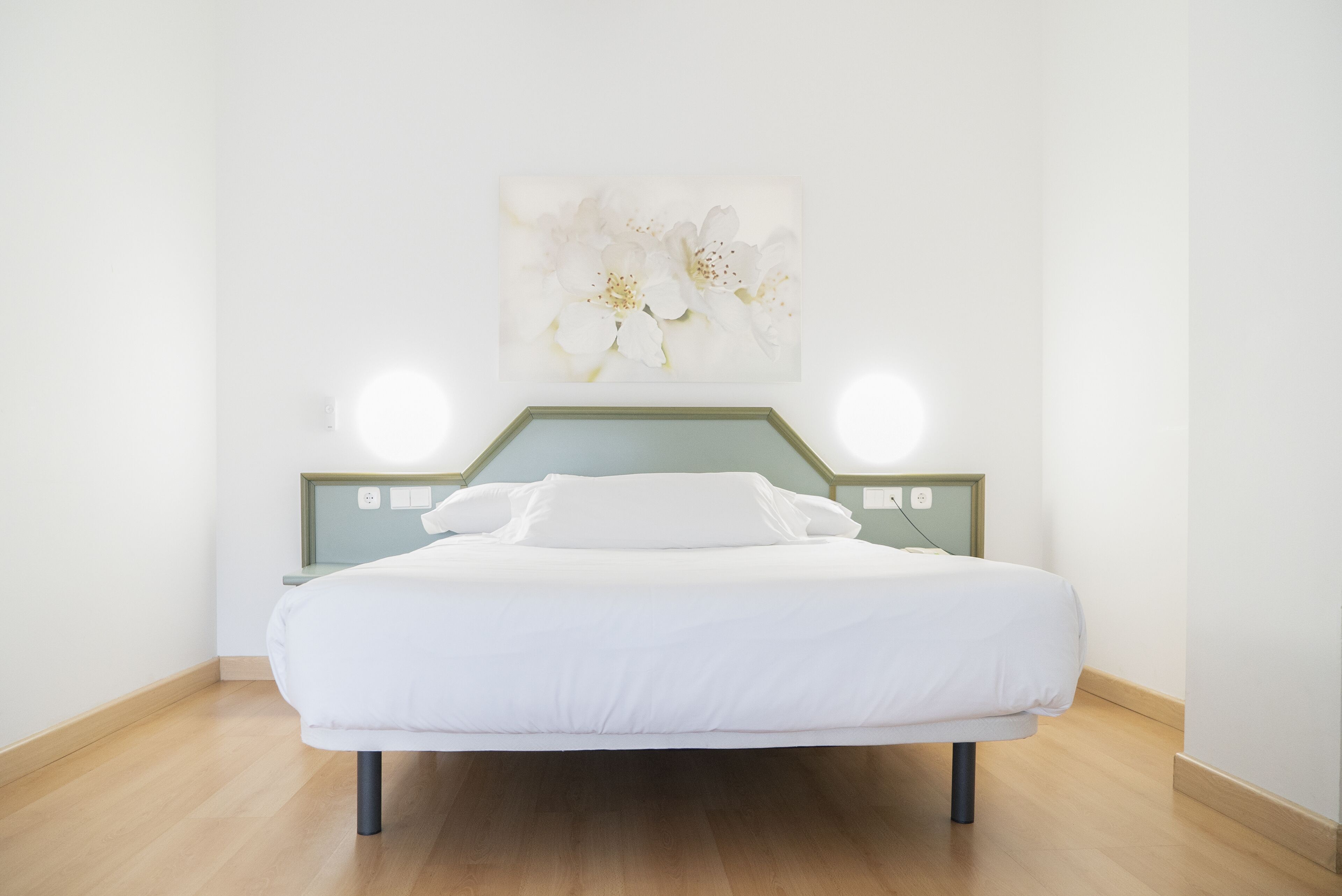 Uma cama bem arrumada com colchão e travesseiros brancos, cabeceira verde e luminária fixada na parede.