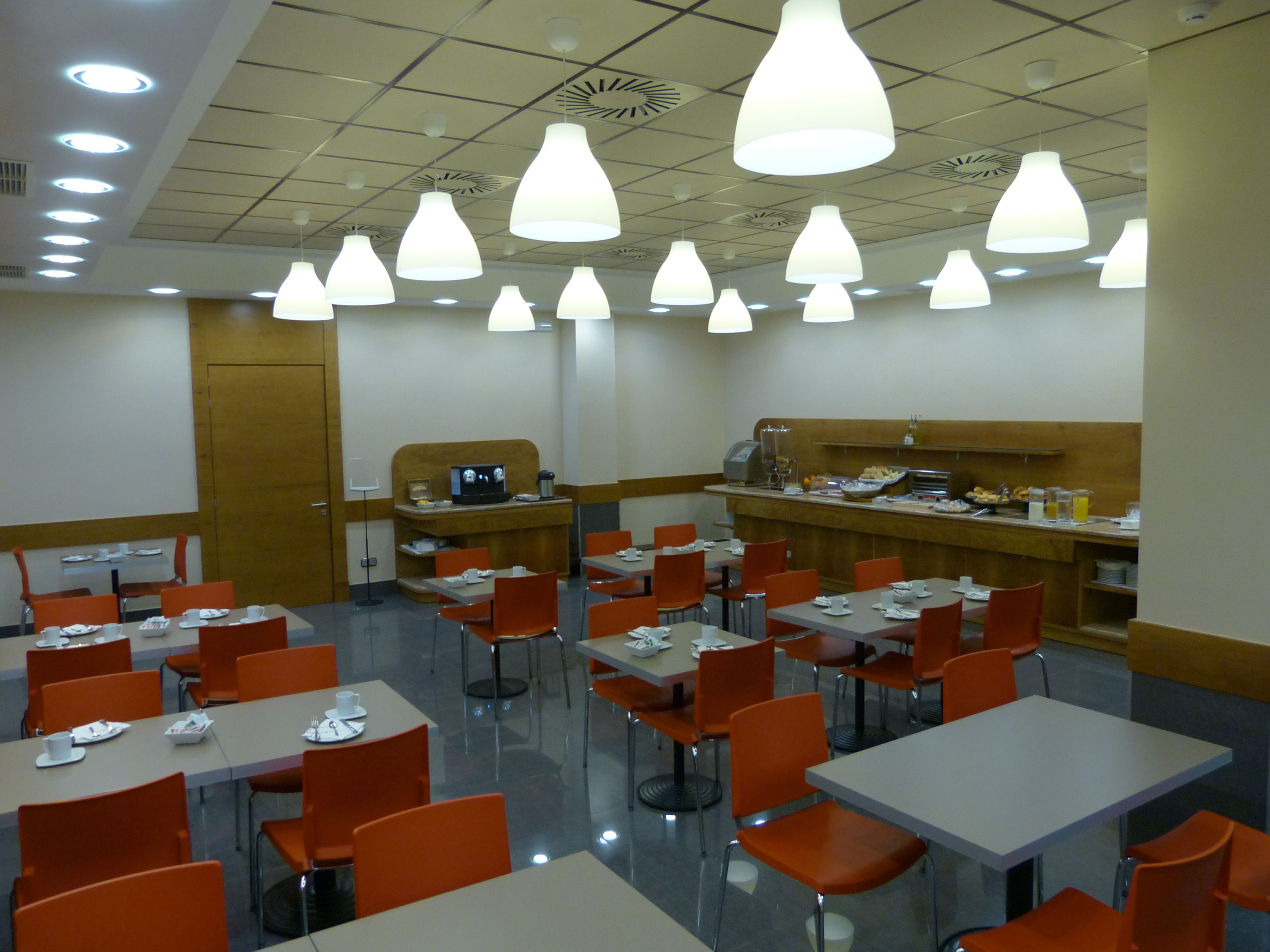 Uma sala de jantar com várias mesas e cadeiras, um balcão de buffet e luminárias no teto.