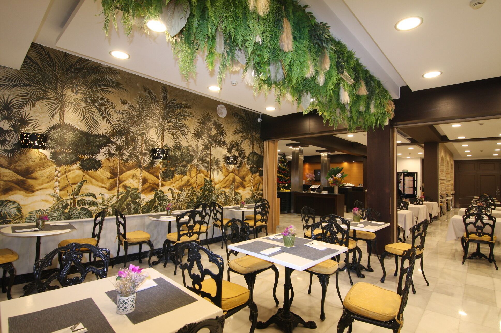 Um restaurante com mesas e cadeiras, um mural com plantas tropicais e um pinheiro de Natal.
